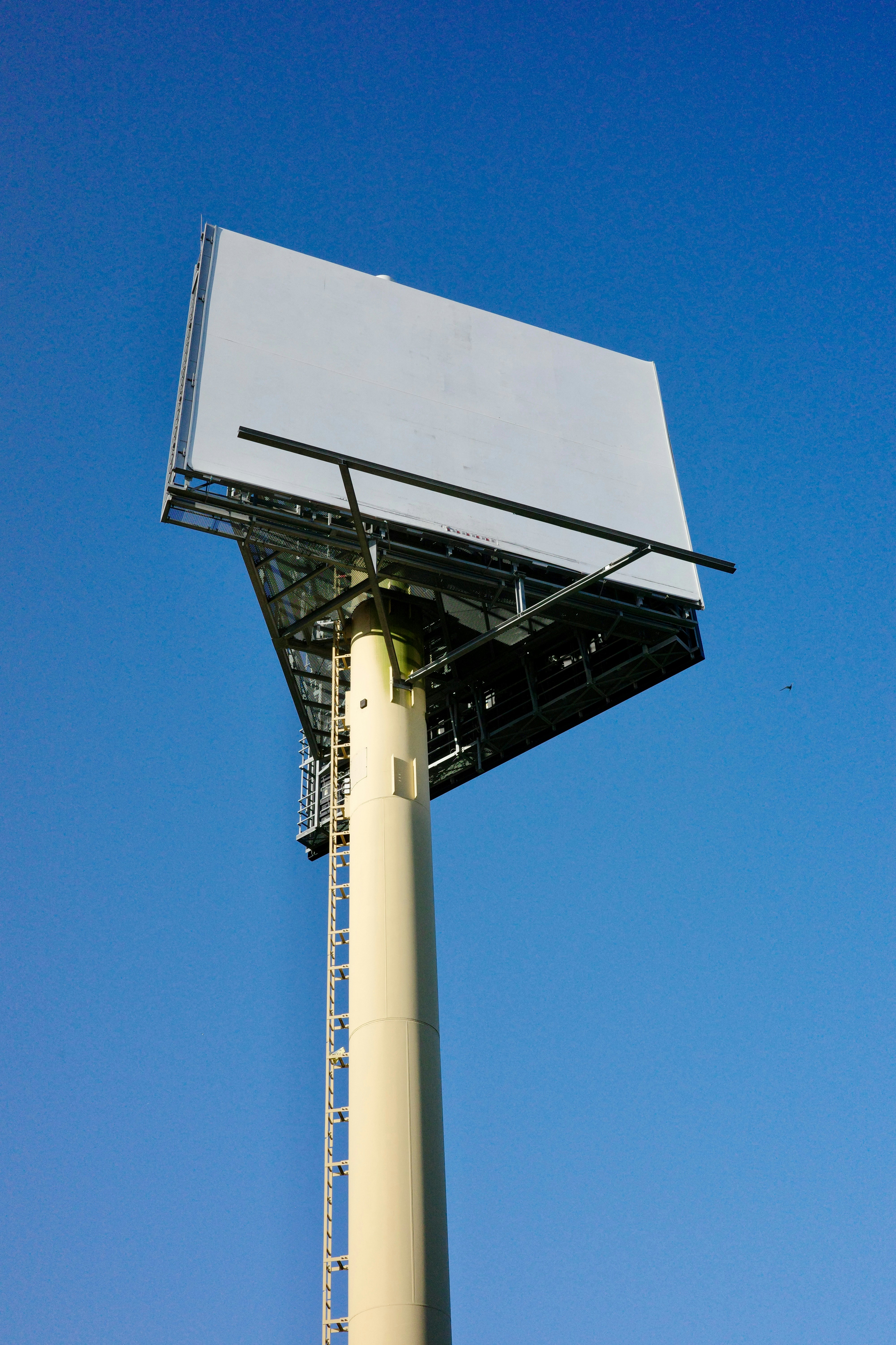 Billboard structure atop a tall beige pillar against a bright blue sky; a stark, minimal composition focusing on industrial geometry and negative space.