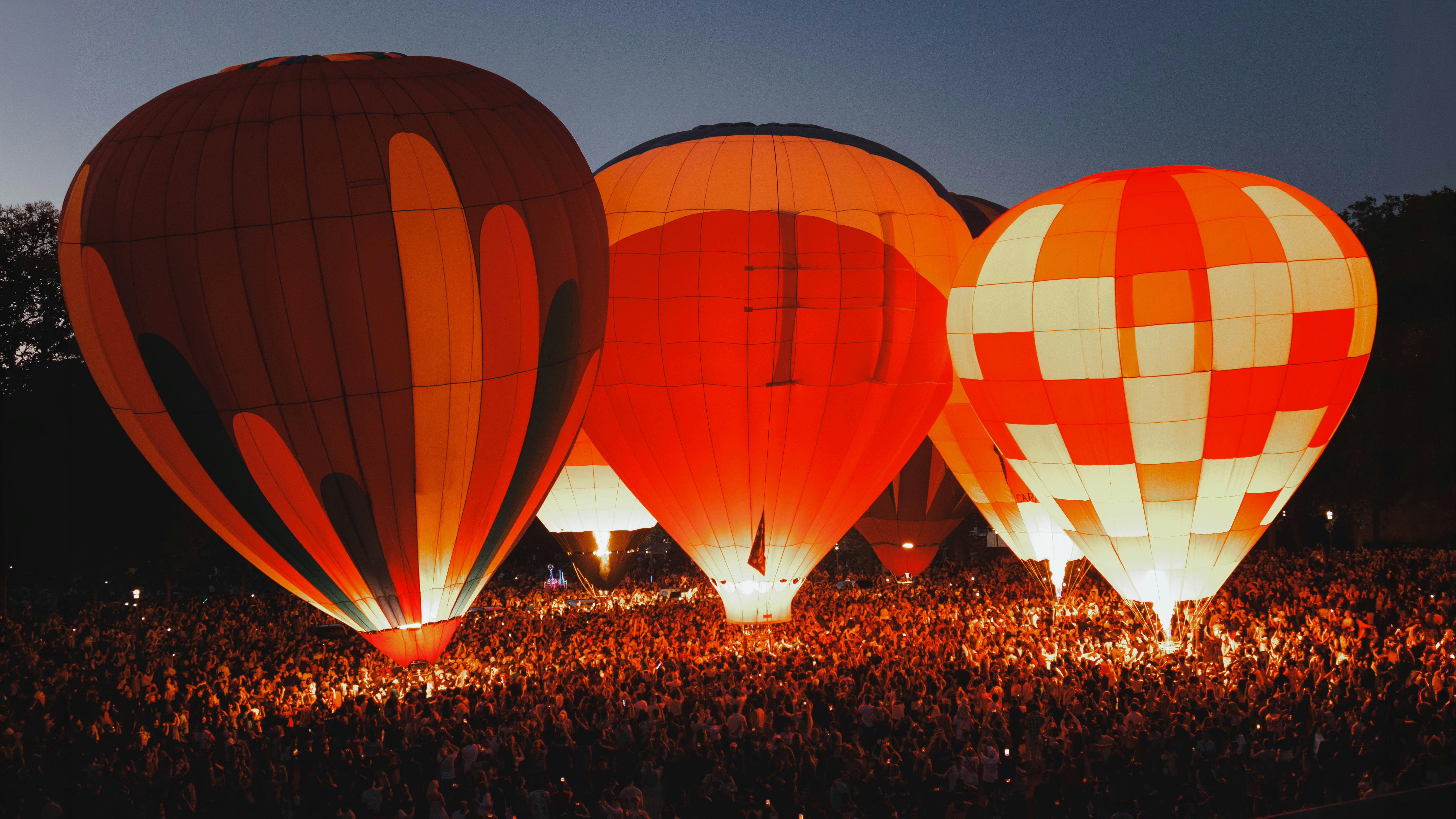 Bright hot air balloons rise above a crowd at dusk, illuminating a packed field.