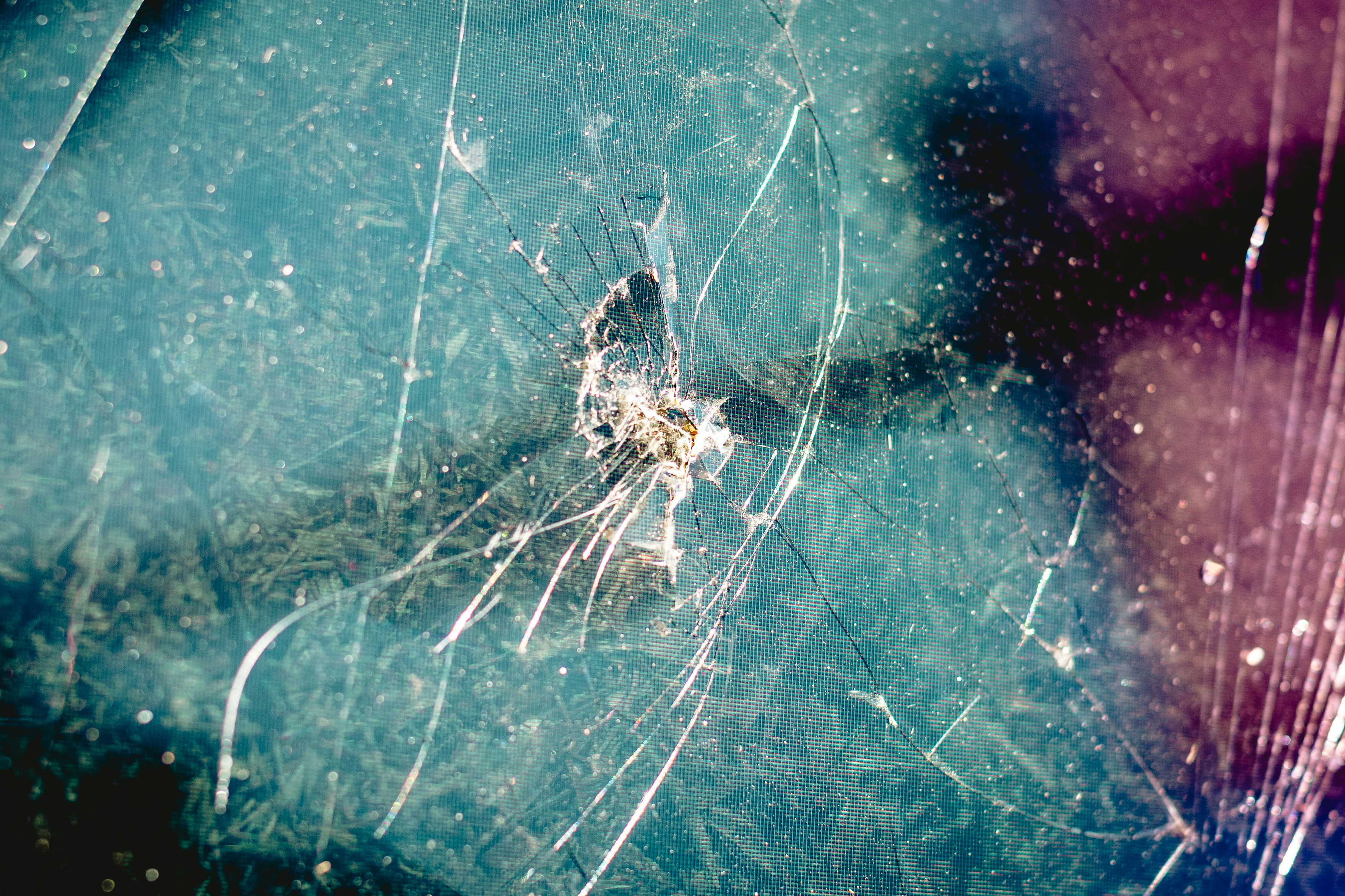 Fractured glass with a central impact mark and light streaks across a tinted background; a concrete abstract close-up suitable for texture or macro study.
