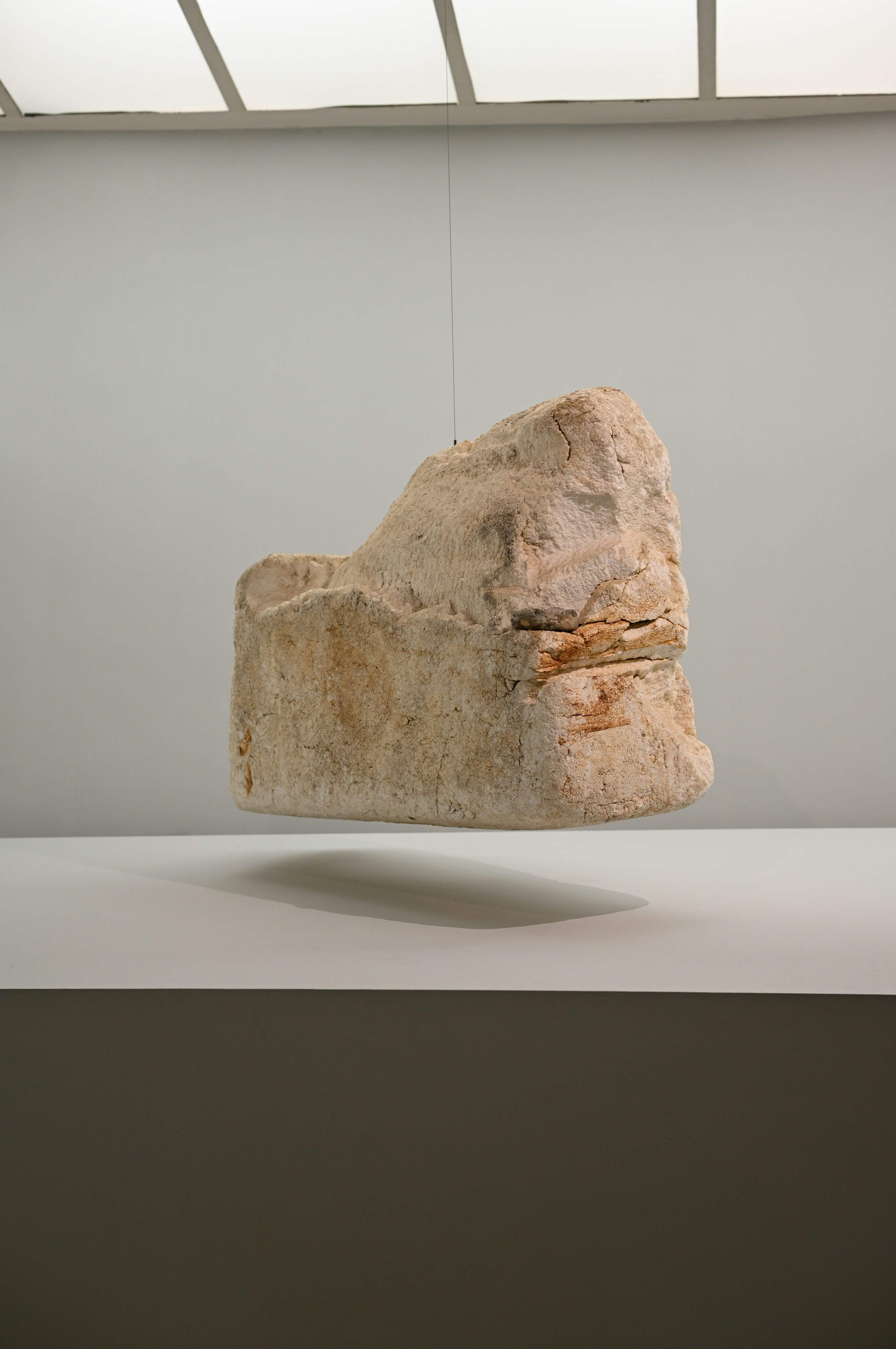 A pale, irregular rock suspended by a thin thread in a minimalist gallery, casting a soft shadow on a white display pedestal.