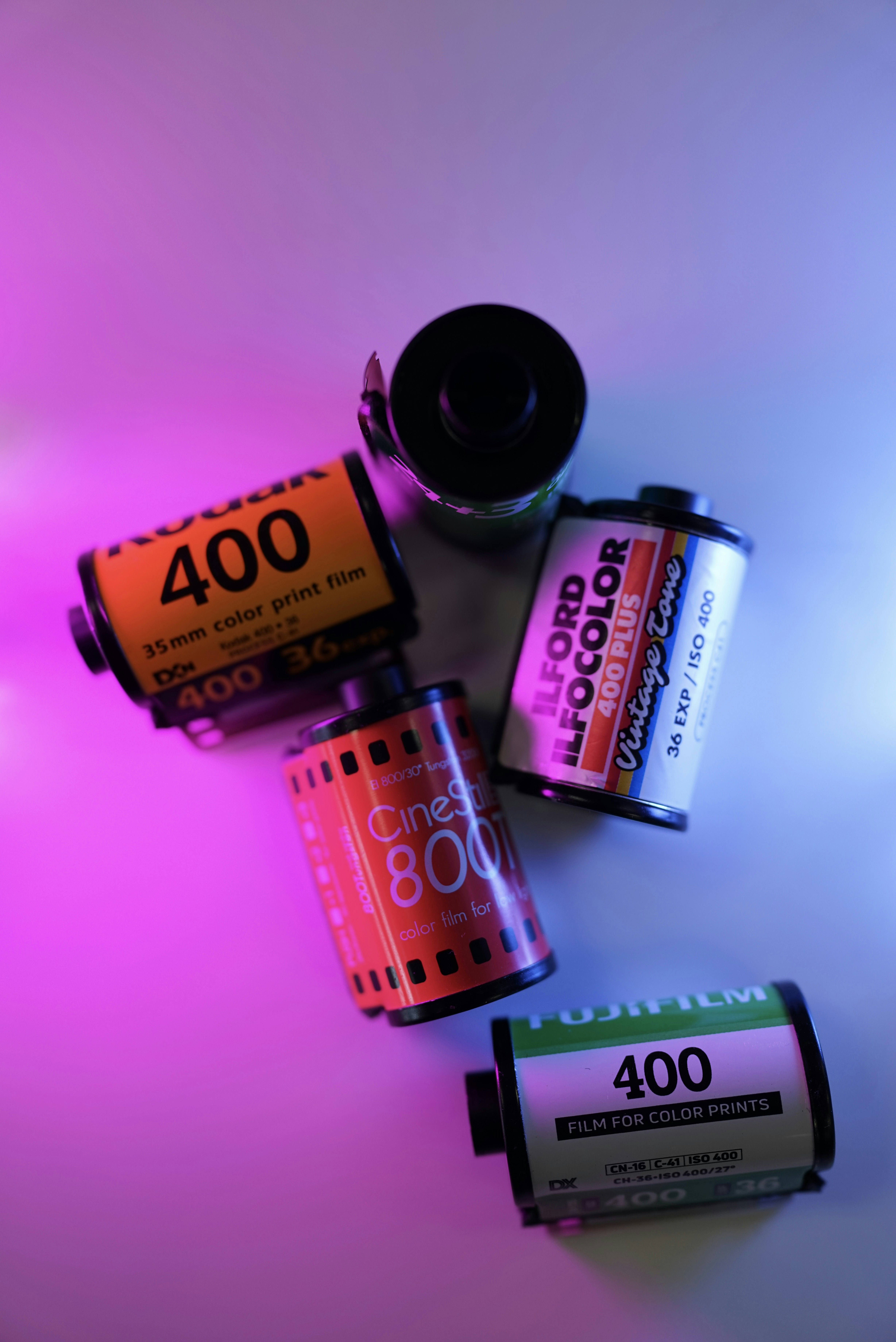 A cluster of vintage color film canisters arranged in a loose circle, illuminated by magenta and cyan lighting, showcasing Kodak and Ilford brands. This is a stylized product/still-life shot of film stock.