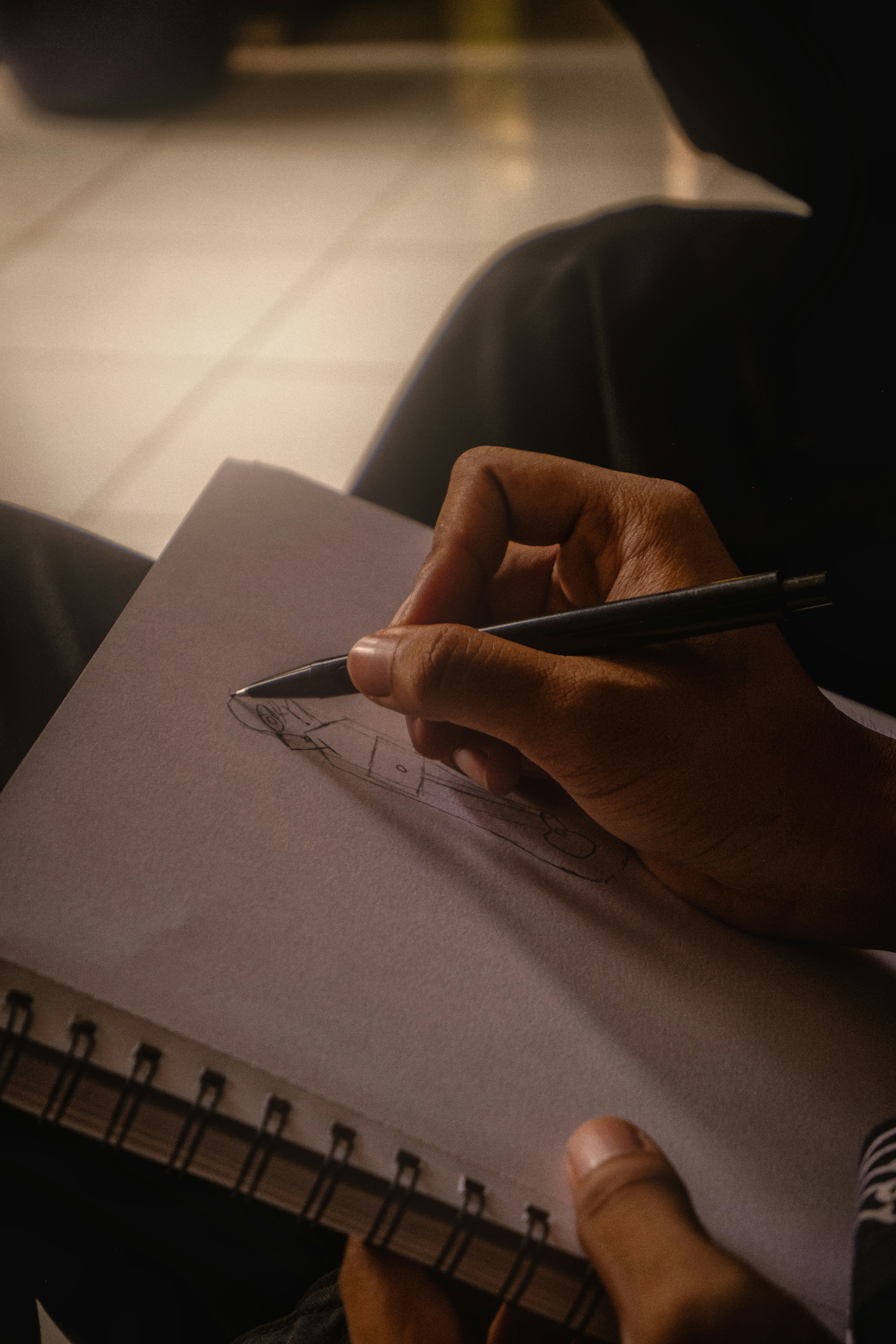 A close-up of a hand holding a pen and sketching in a spiral notebook, with soft ambient lighting and blurred background.