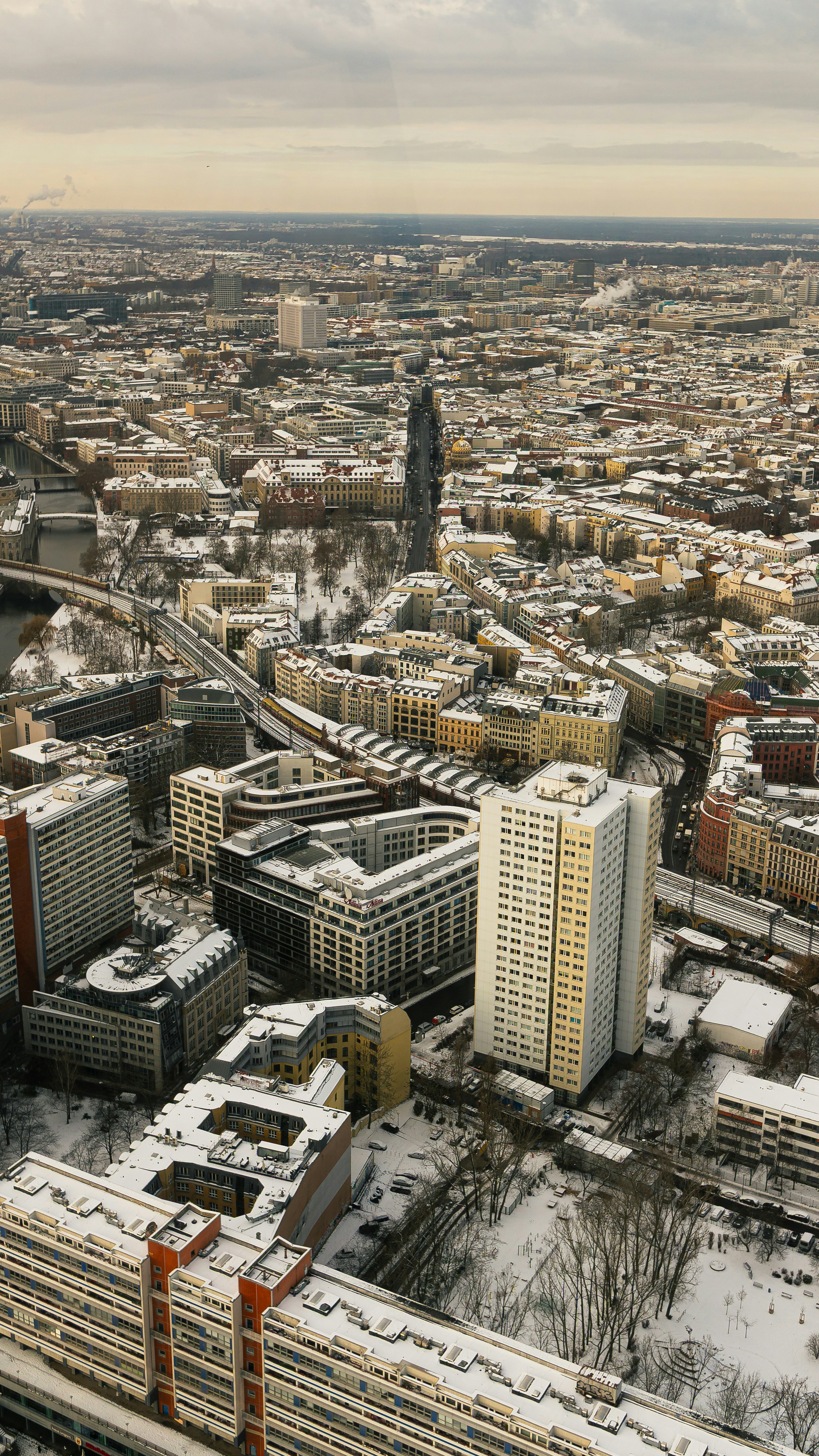 Aerial view of a dense urban landscape dusted in snow, with high-rise and mid-rise buildings, streets carving through the grid, and faint smoke plumes rising in the distance. Snow emphasizes rooflines and avenues below.