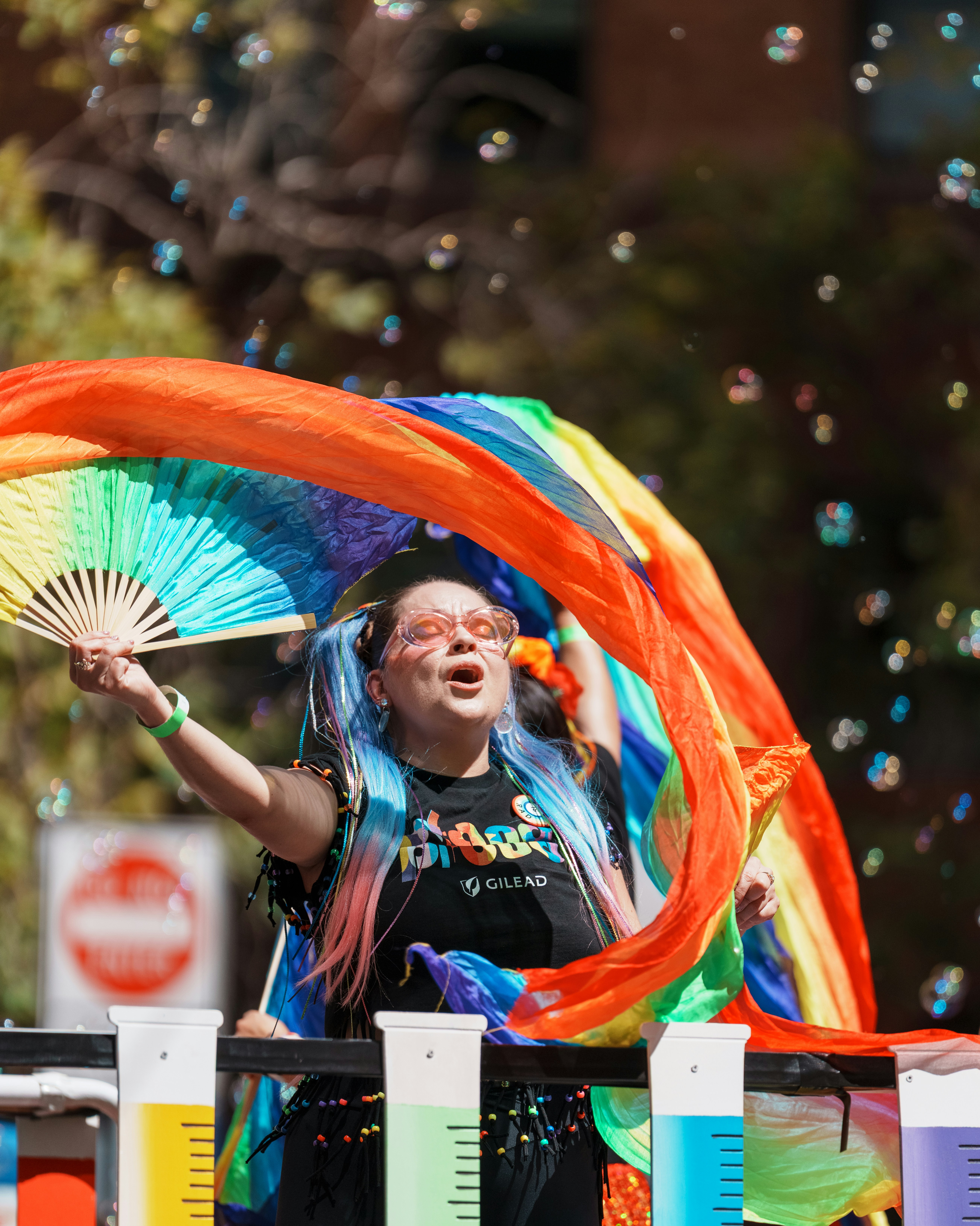 Vibrant performer waves a rainbow scarf and handheld fan during a lively outdoor celebration, with confetti-like bubbles and a cheering expression in focus.