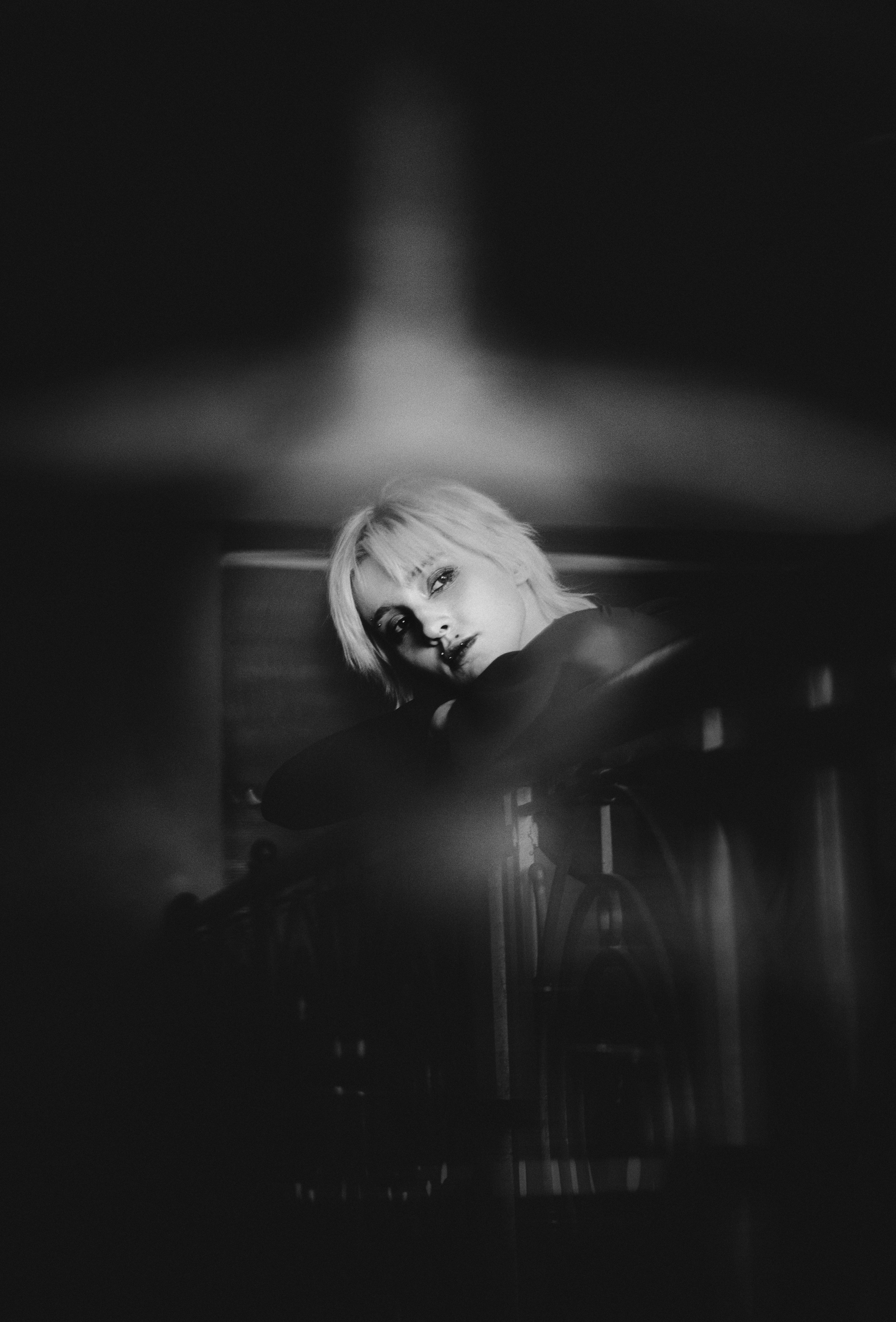 A monochrome portrait of a woman with bleached short hair leaning on a railing on a dim balcony, soft focus and natural vignette creating a moody, intimate mood.