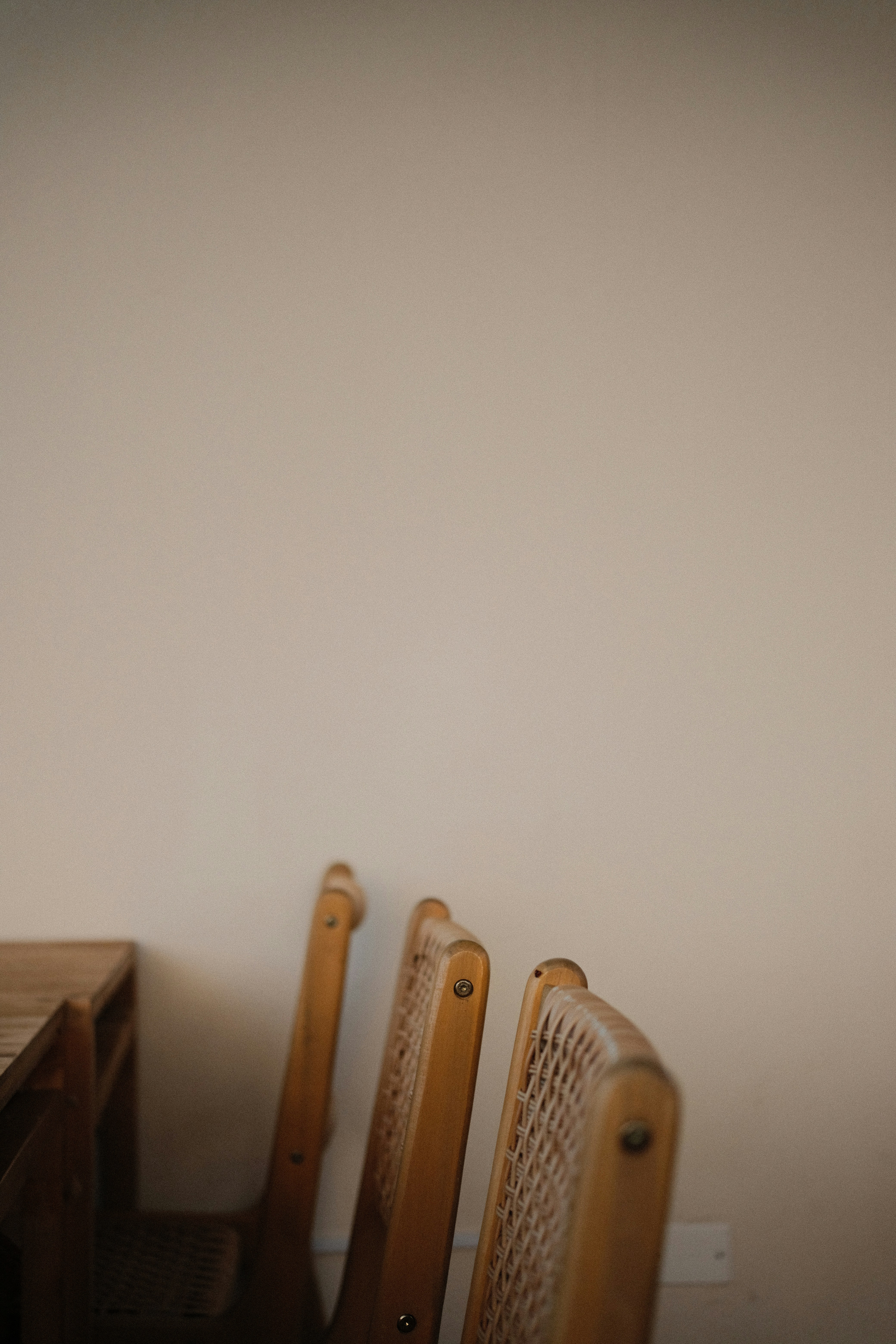Simple interior scene featuring a row of wooden chairs leaning against a pale wall, with a desk edge visible to the left. The composition emphasizes negative space and subdued tones.