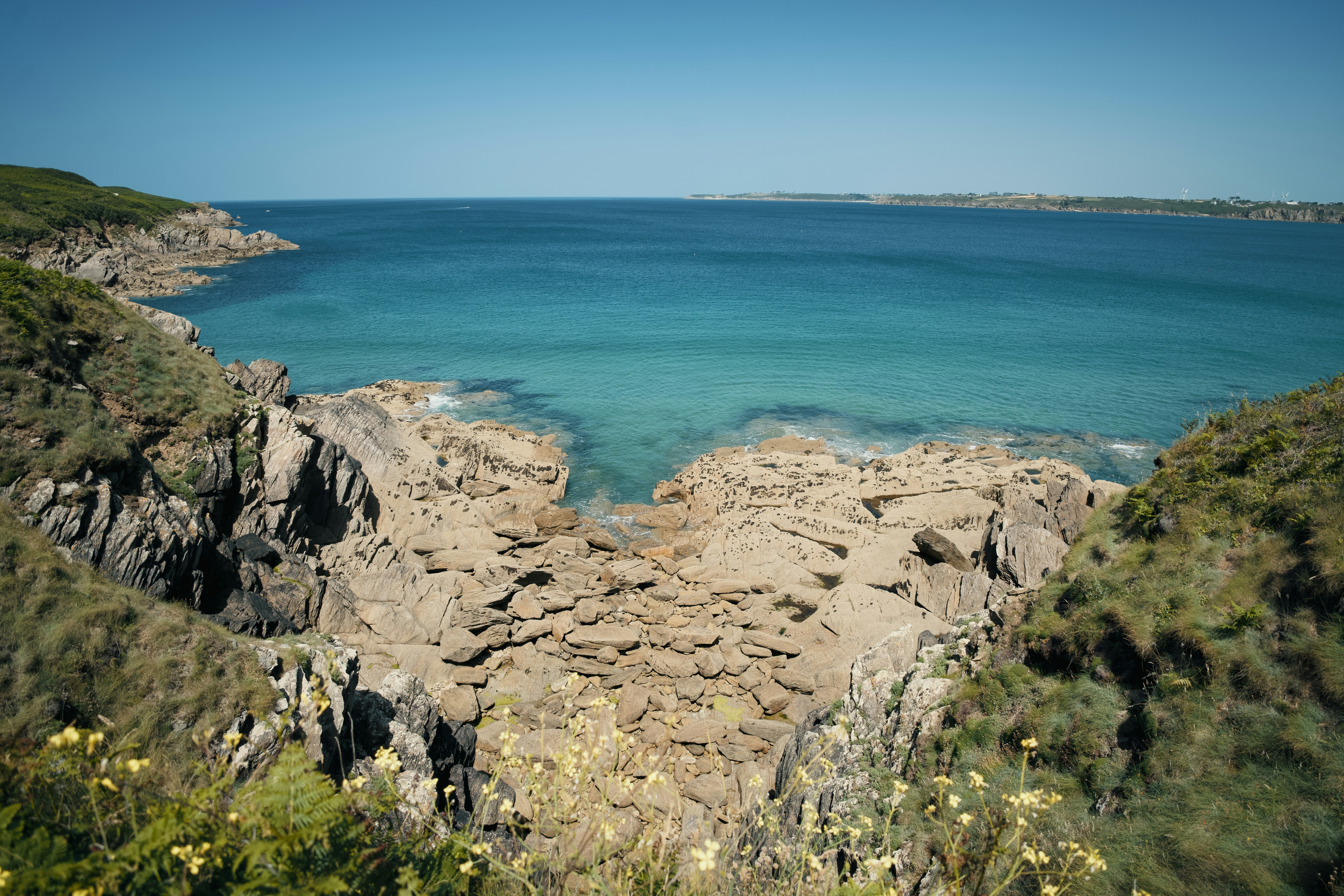 Sunlit coastal rocks frame a turquoise sea with a distant horizon; foreground shows rugged terrain and sparse yellow wildflowers, capturing a tranquil shoreline overlook.