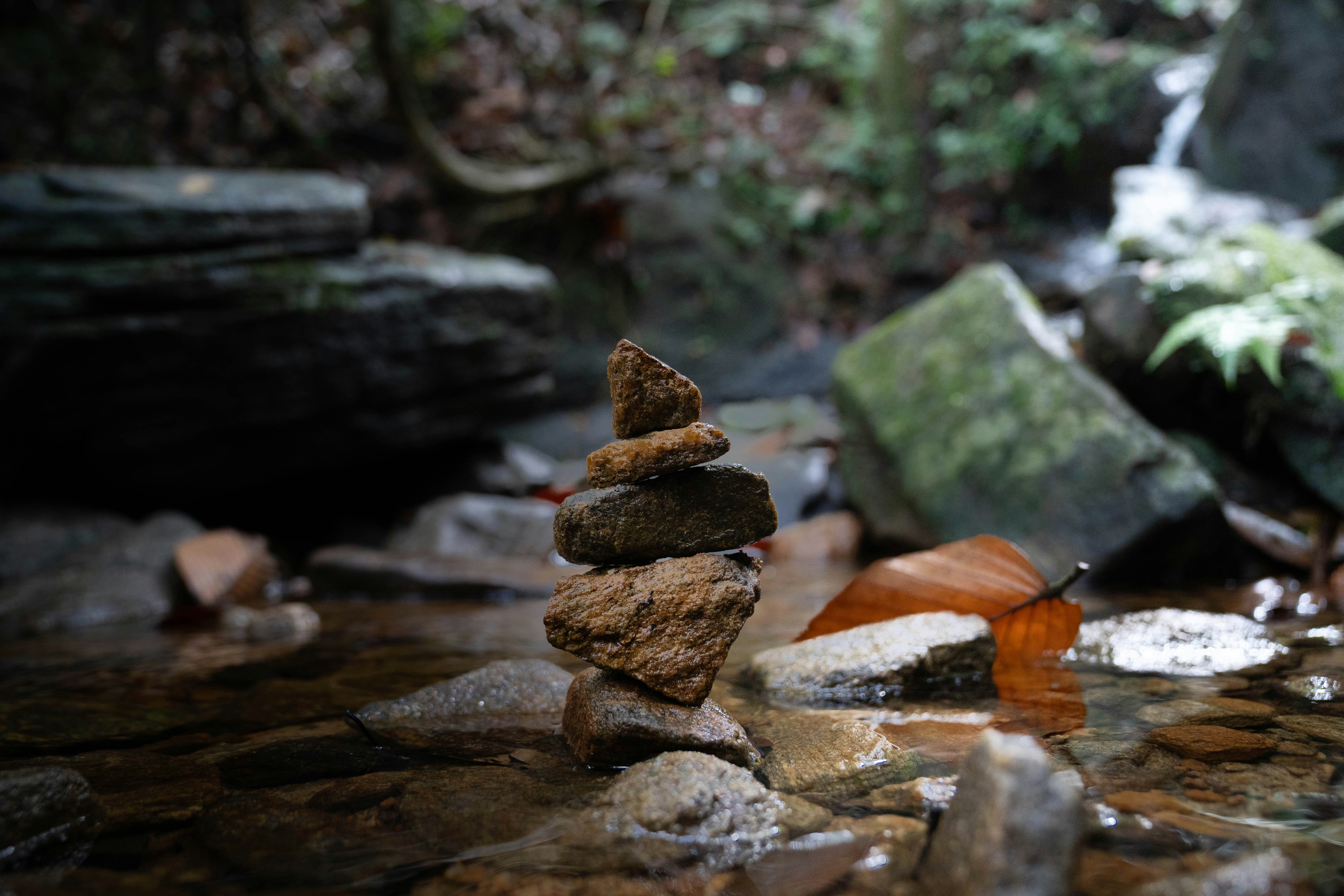 Stacked stones balance in a shallow stream with a blurred forest backdrop and a hint of a waterfall; a calm, nature-focused scene with a rocky foreground.