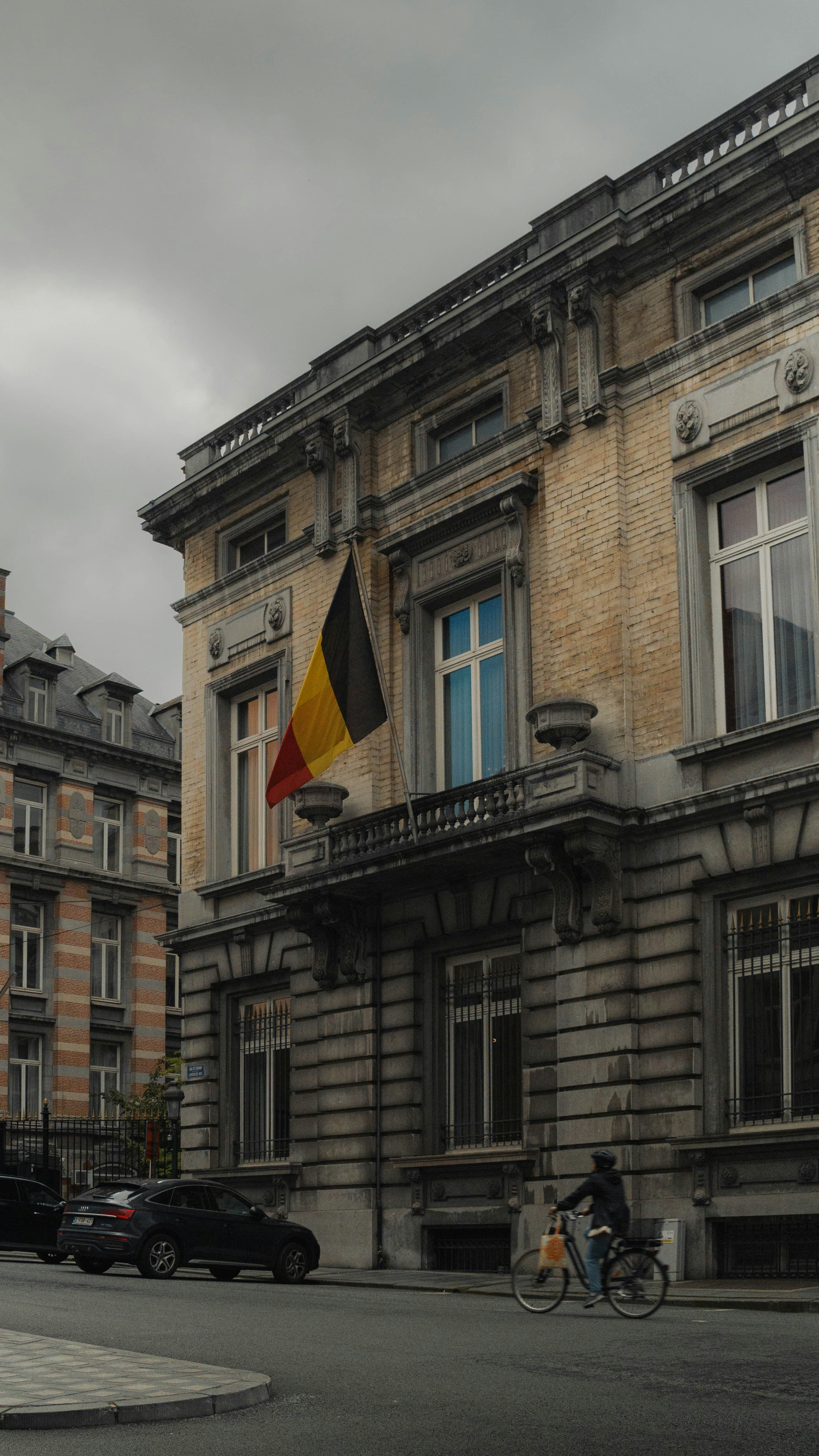 Belgian flag on ornate building with cyclist on urban street.