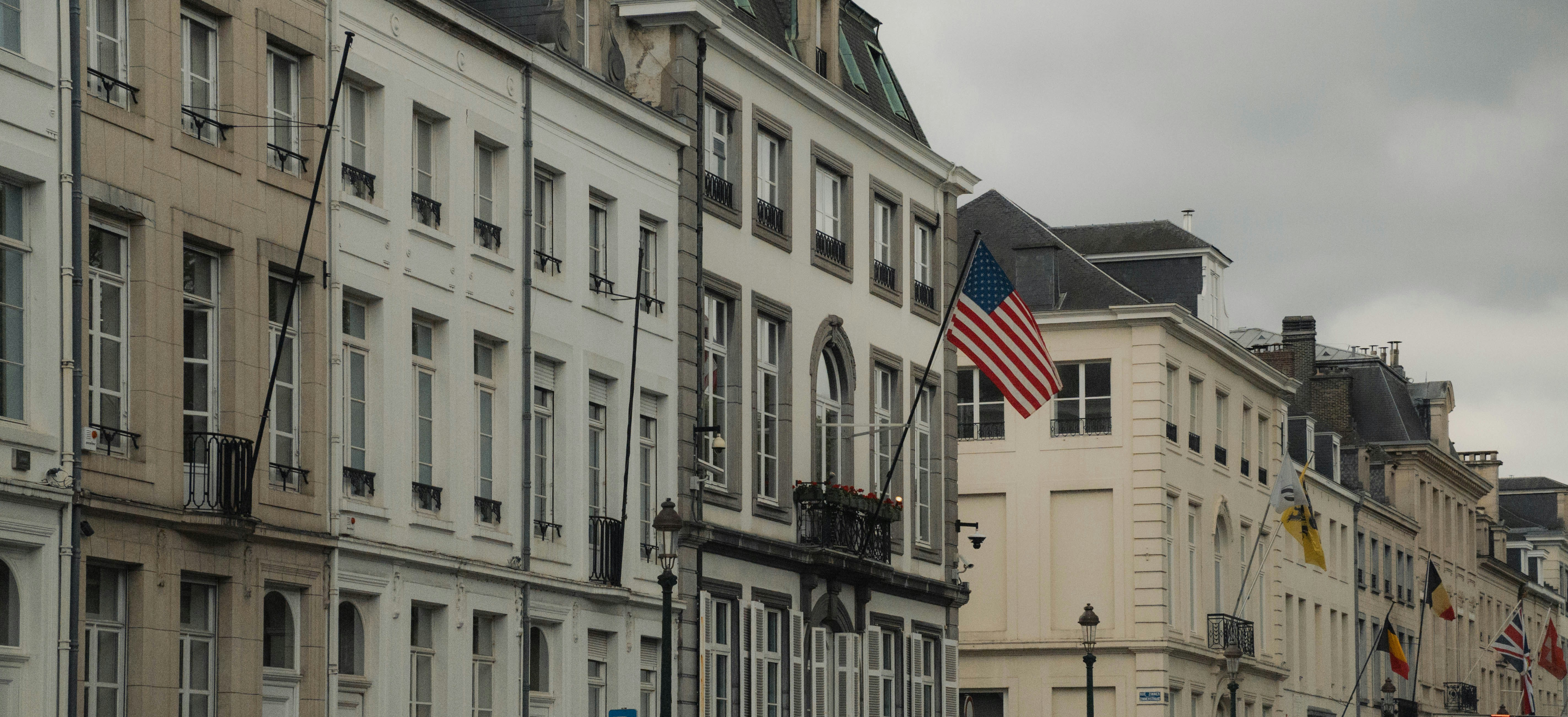 Row of beige and white historic townhouses with multiple flags along a quiet street; American flag prominently displayed, overcast sky hinting at a calm urban morning.