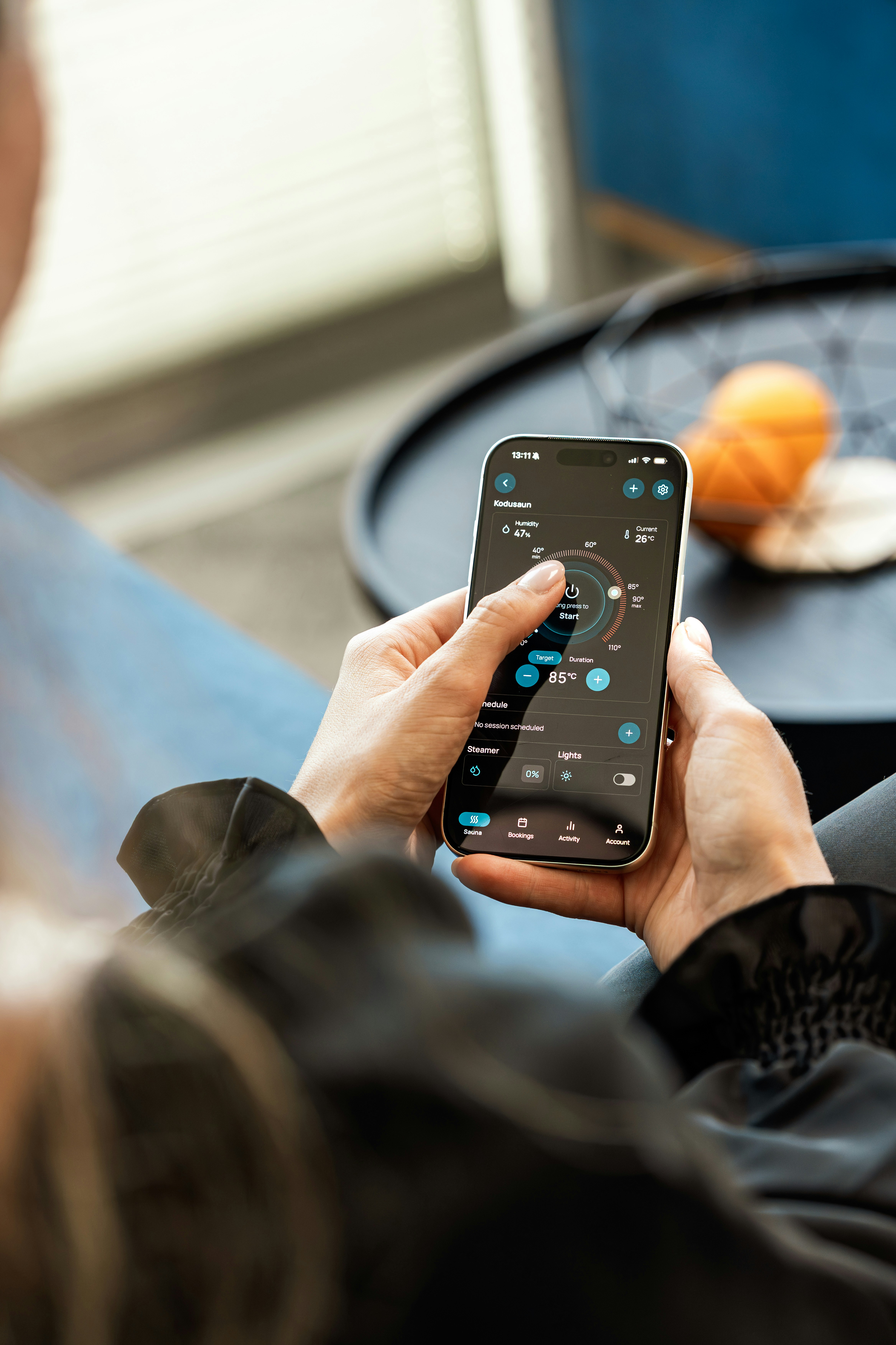 A person holds a smartphone displaying a detailed control interface in a cozy indoor setting with a table and orange decor in the background.