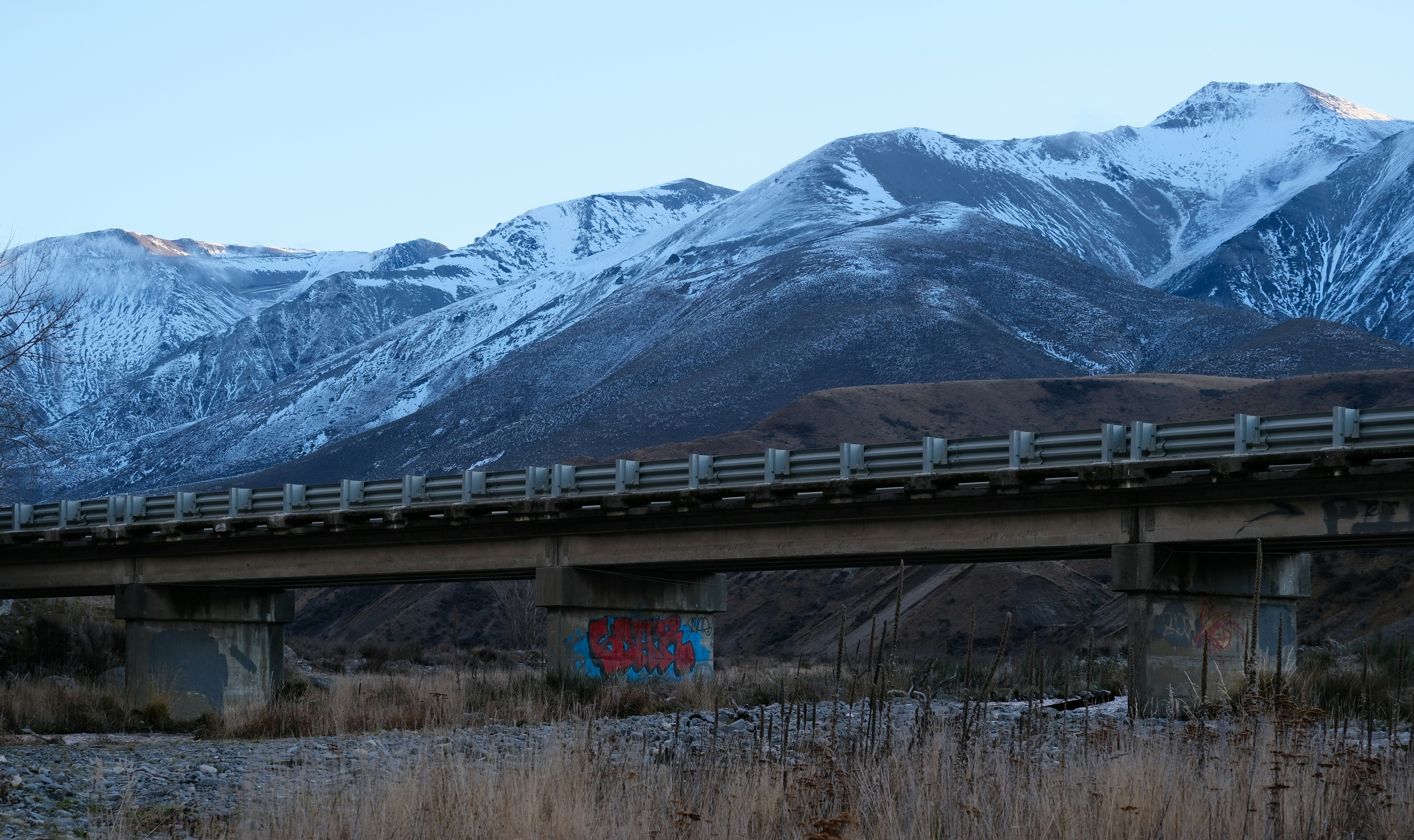 Bridge spanning a rocky riverbed with snow-dusted mountains in the background, set under clear light; the scene emphasizes rugged terrain and engineering against a alpine landscape.