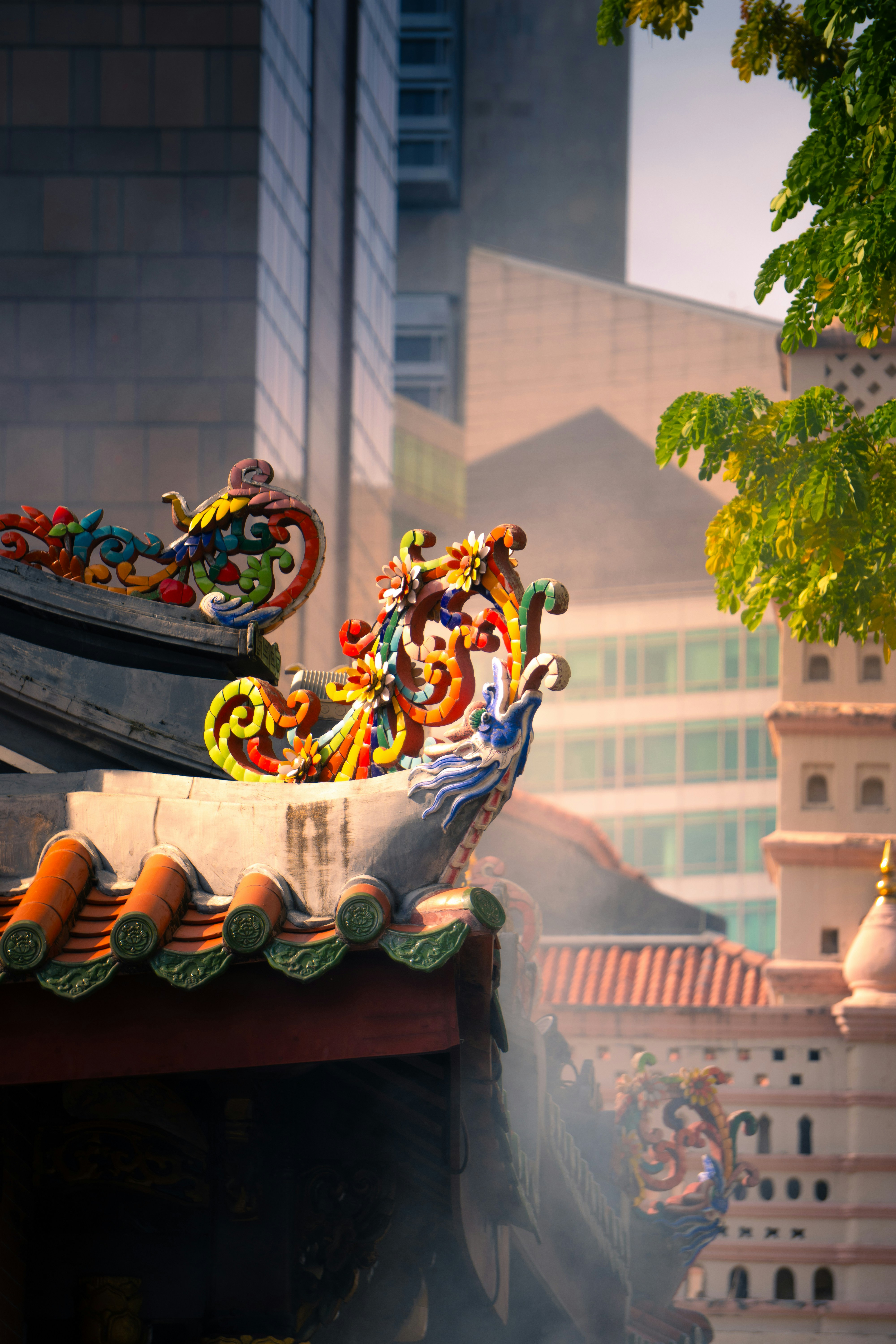 Colorful temple rooftop carvings framed by urban architecture and green foliage, revealing cultural detail amid modern buildings.