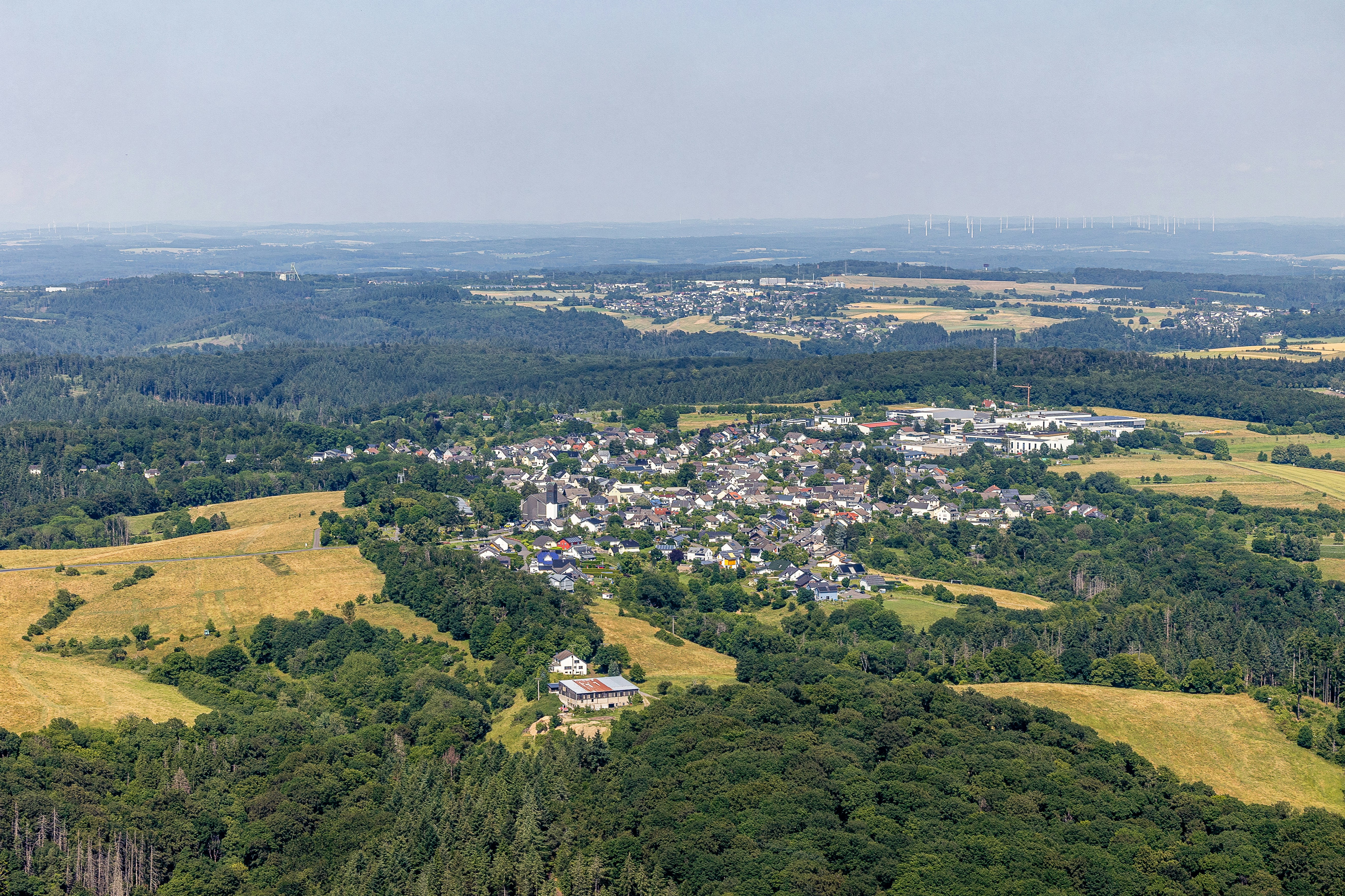 Aerial view of a small village nestled among patchwork fields and forests, captured from above to reveal surrounding rural landscape and distant horizon.