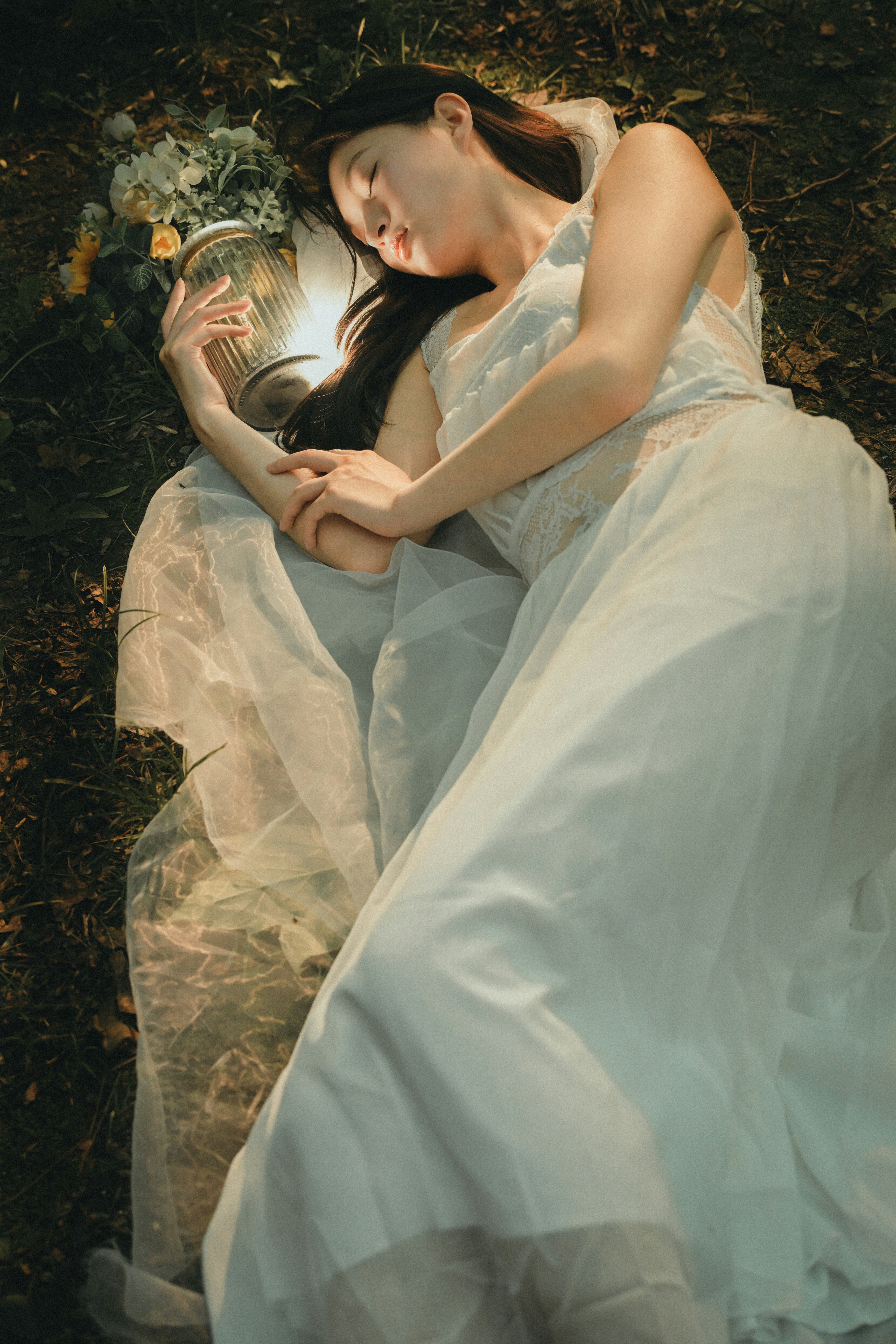 Young woman in white dress lying with glowing lantern outdoors.