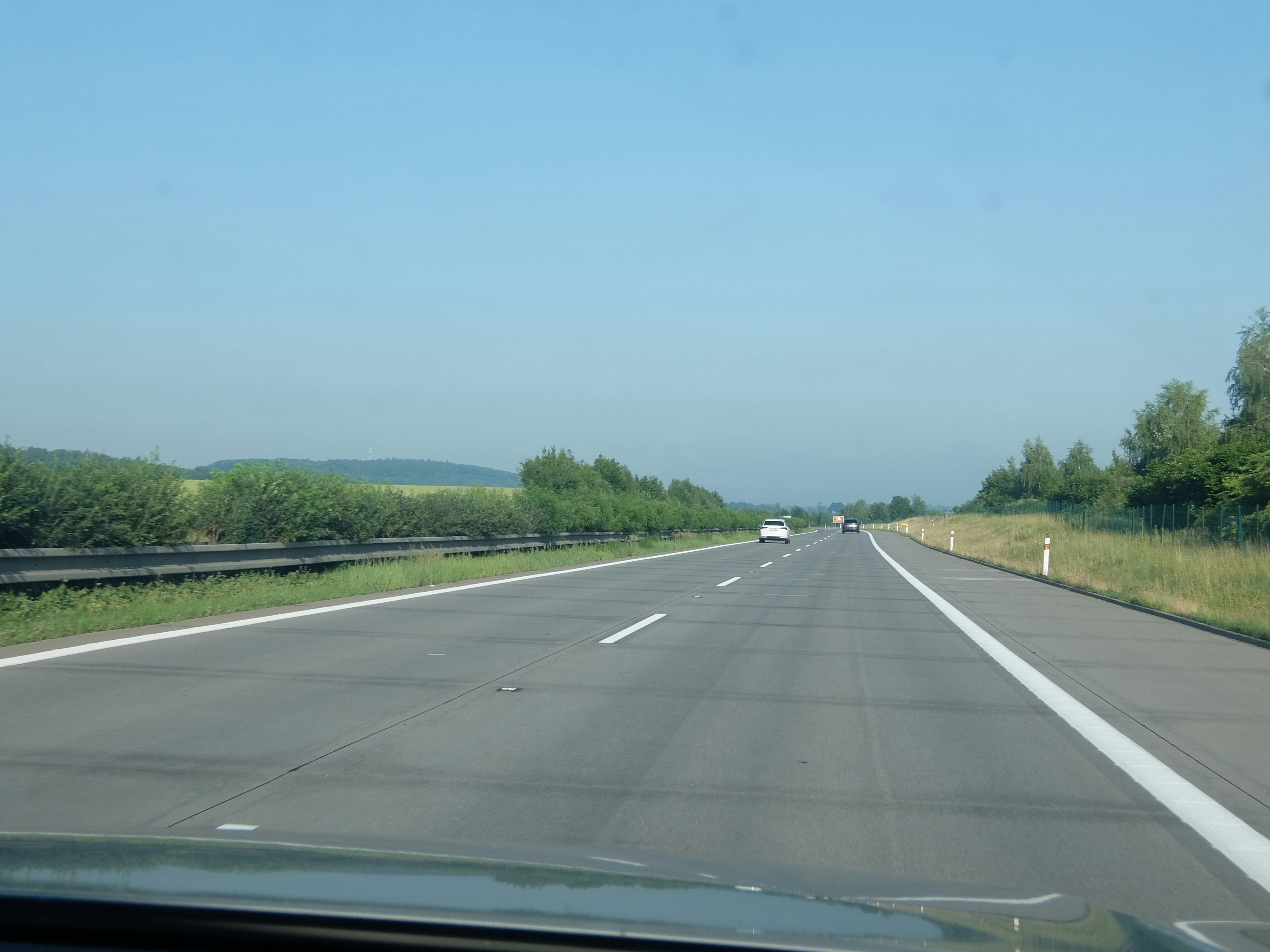 Sunlit highway stretches into the distance with a lone car, flanked by greenery under a clear blue sky.