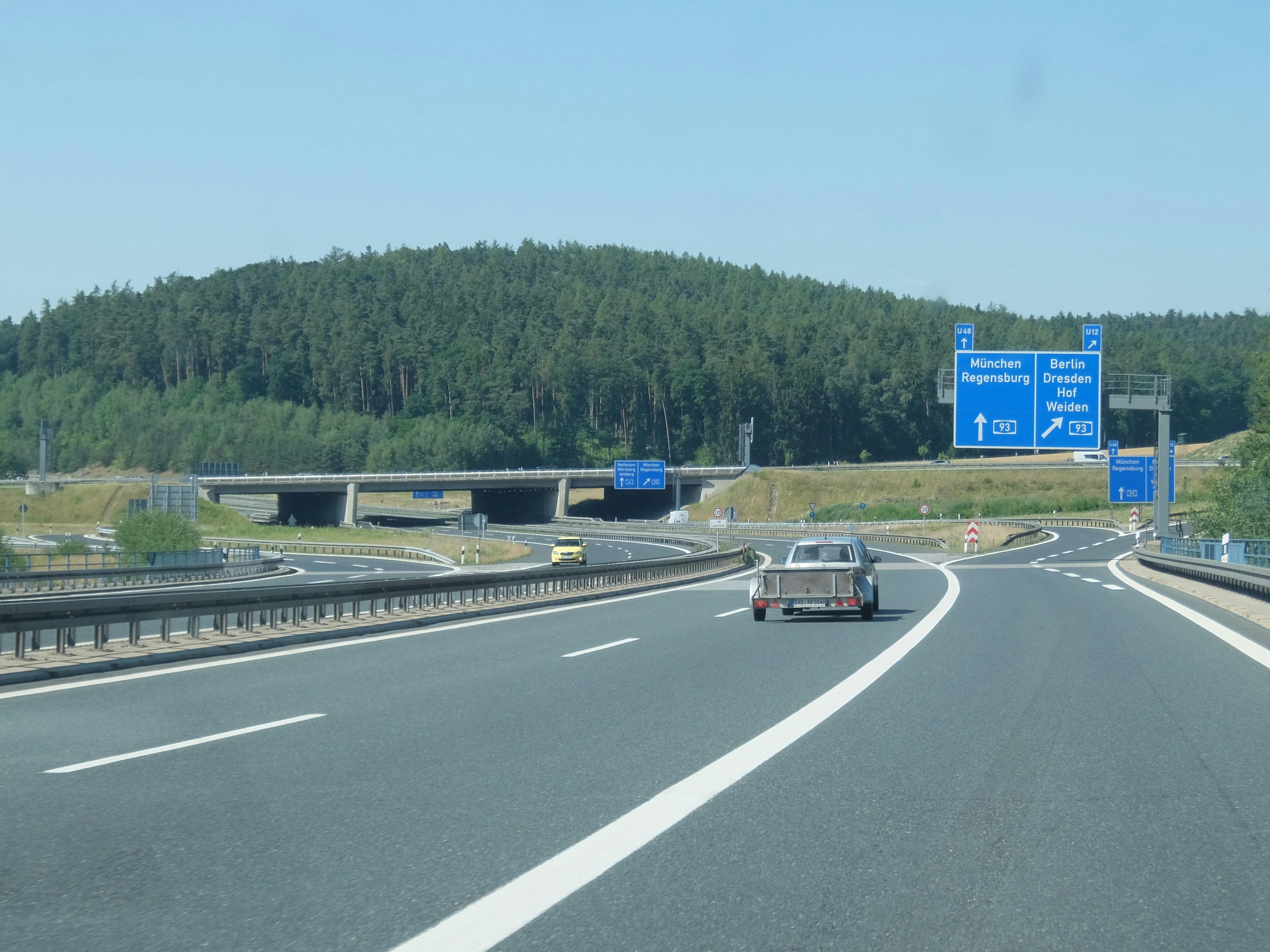 Stretching highway with a distant car under blue sky and a forested horizon; a signposted junction communicates routes ahead, conveying motion and destination.