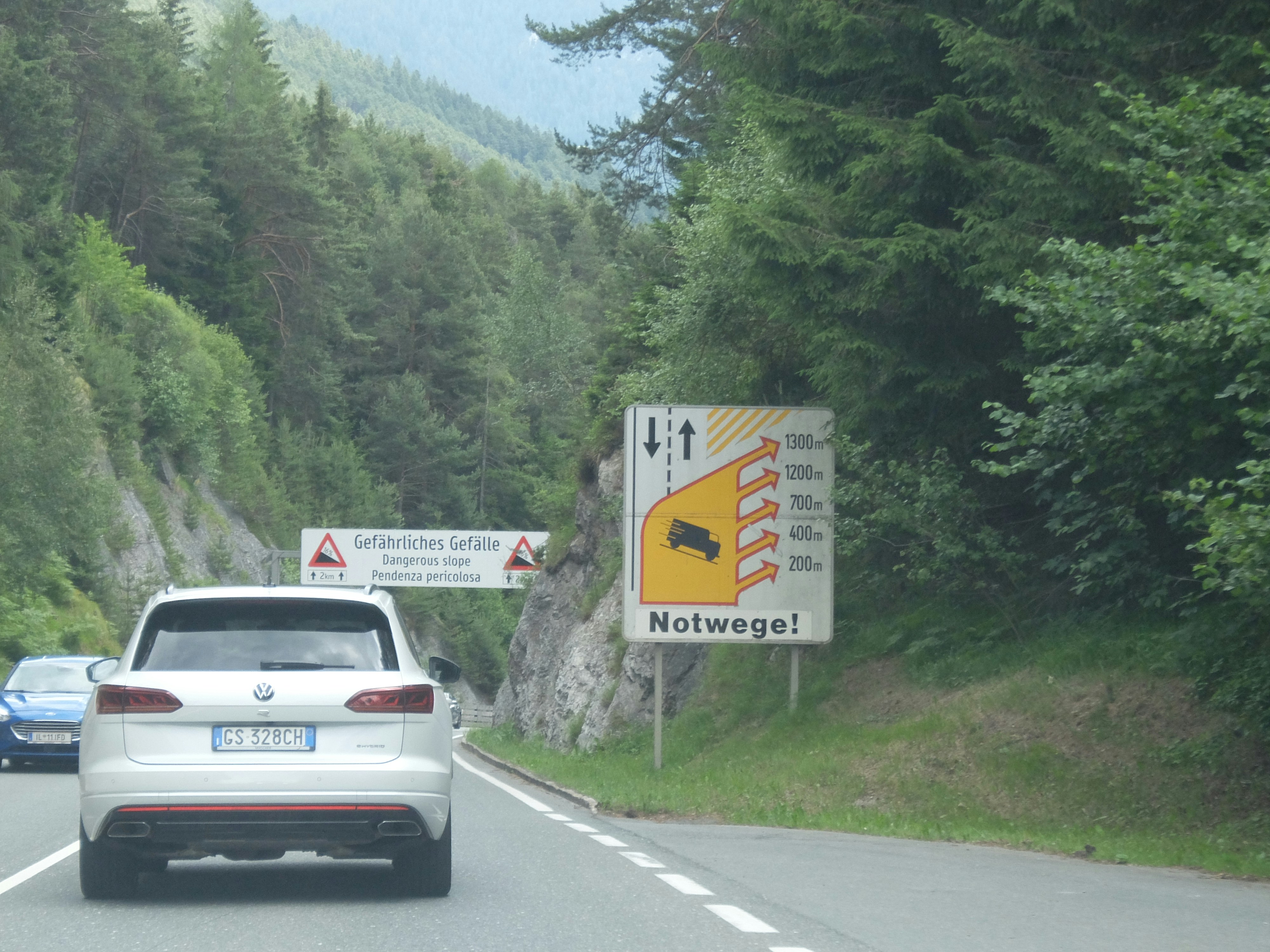 White SUV leads a two-car convoy along a mountain road flanked by evergreen trees and warning signs.