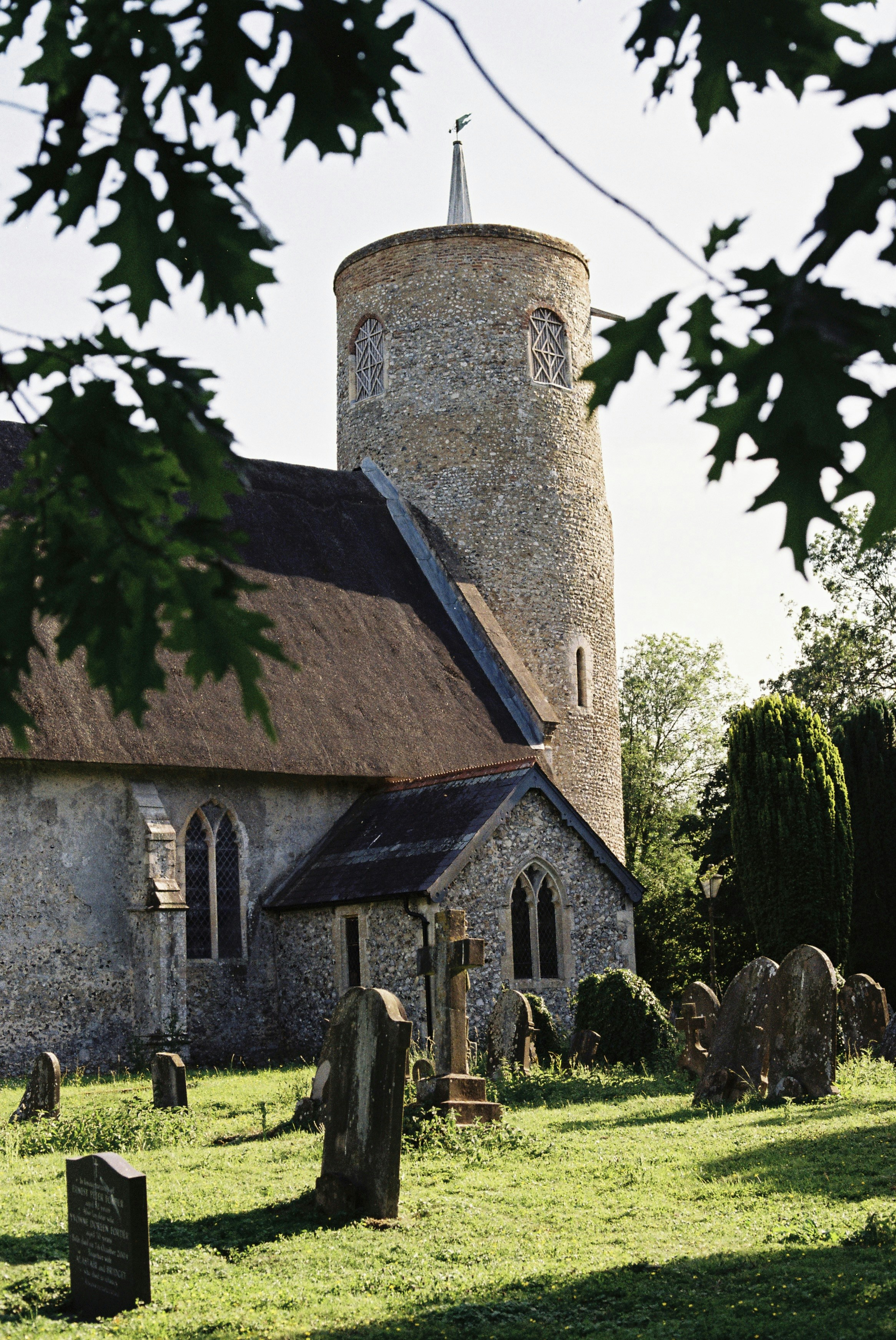 Sunlit stone chapel with a tall circular tower sits amid a grassy graveyard; foreground branches frame the scene with weathered tombstones and a sloping roof.