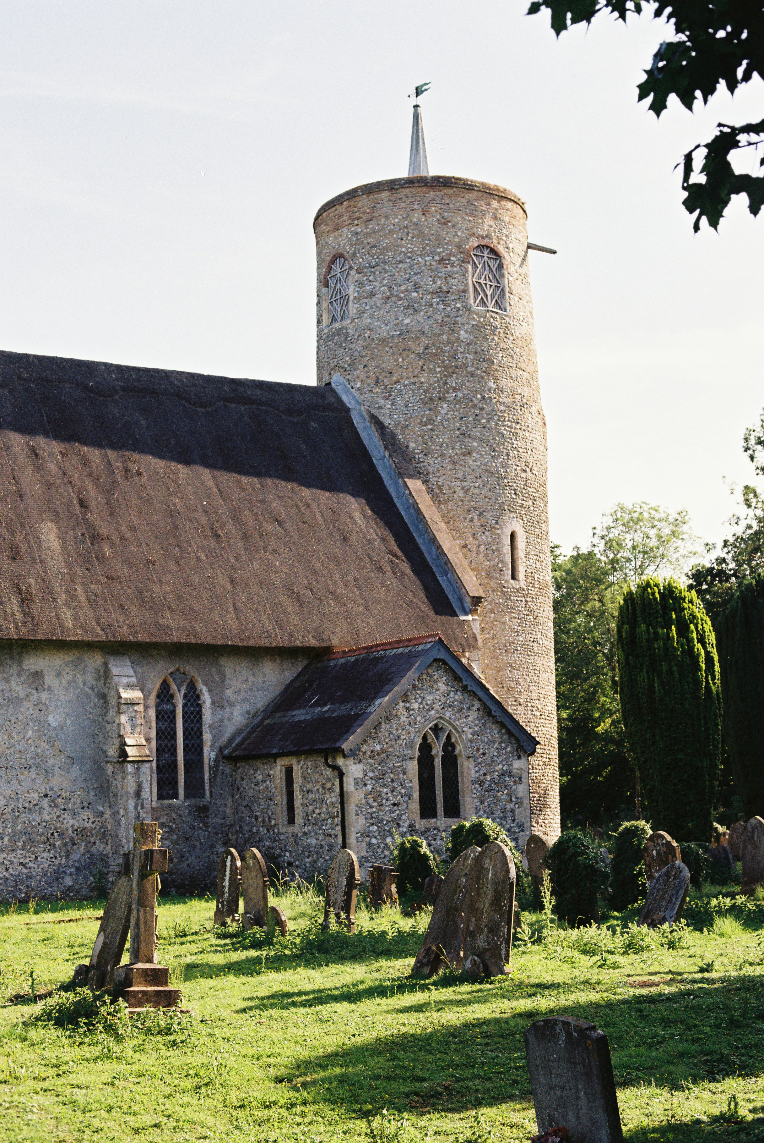Sunlit stone church with a tall circular tower sits among aged gravestones in a quiet churchyard.