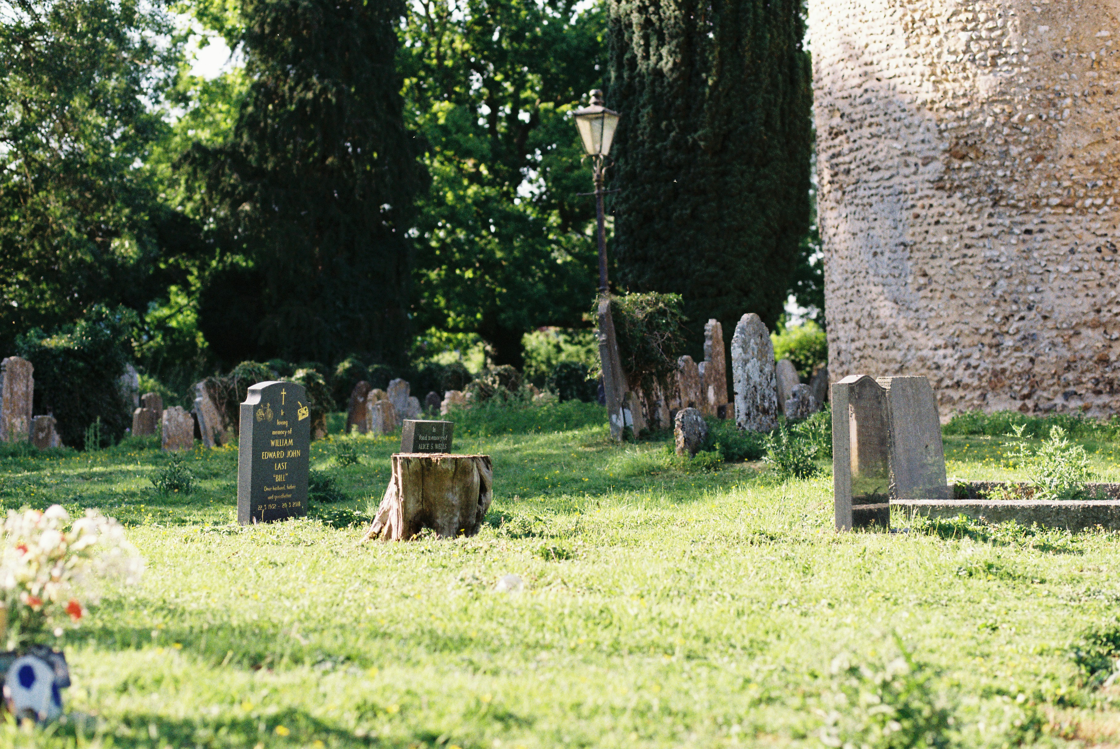 Sunlit cemetery with weathered headstones and a stone tower along a grassy path; a lamppost rises above the scene, conveying tranquil, timeless repose.
