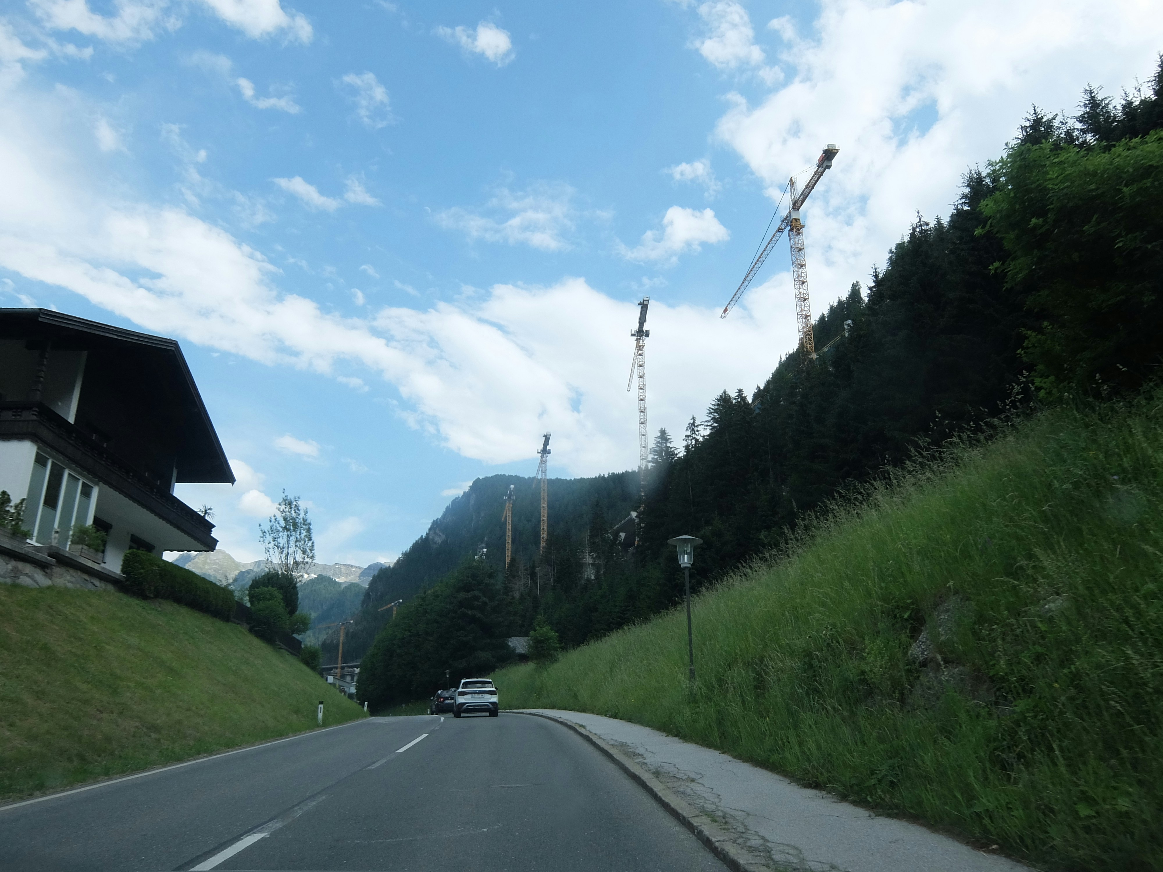 Open highway winding between green hills with construction cranes rising above a distant mountain range.