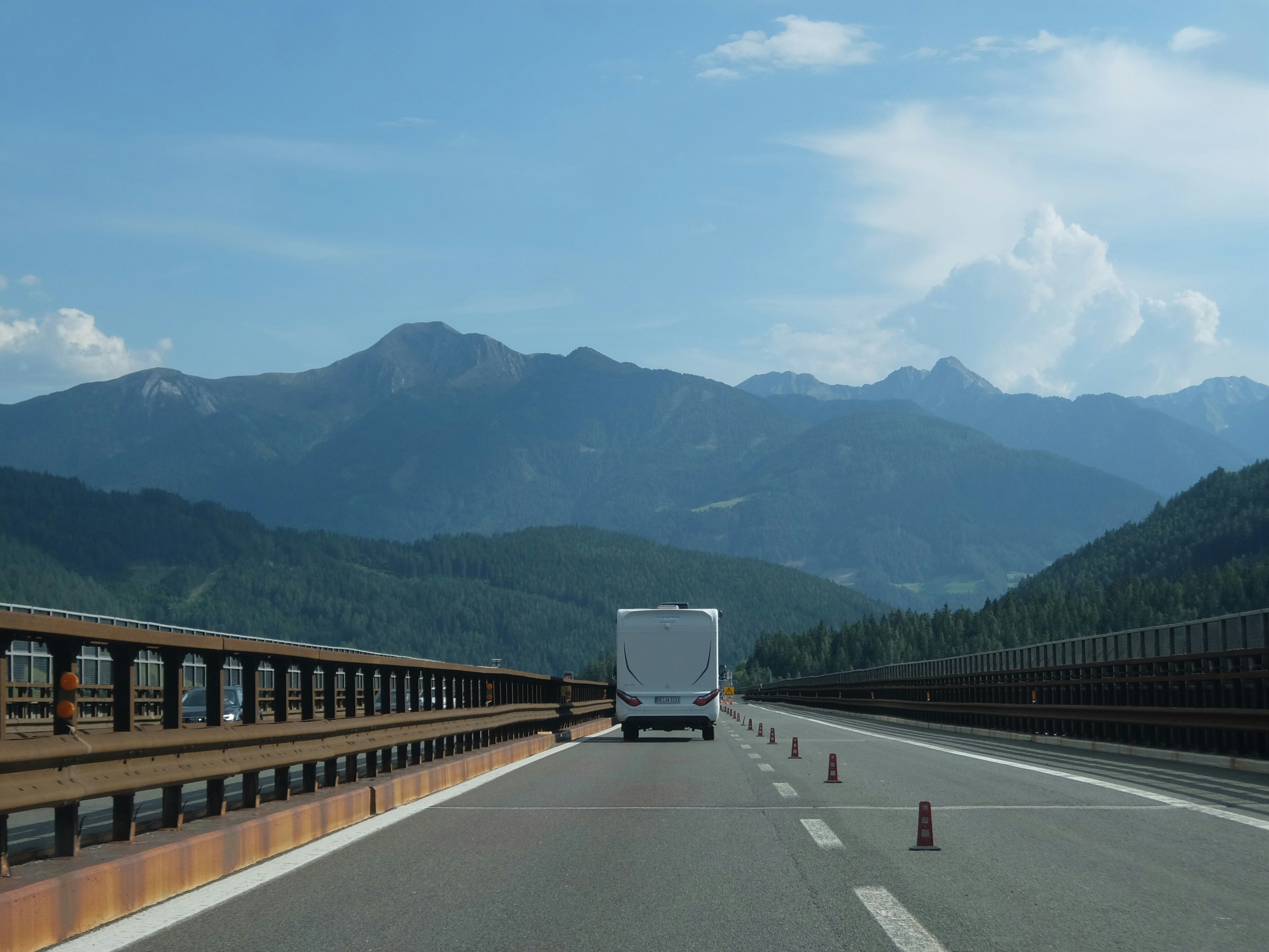 A lone white caravan travels down a long, empty roadway flanked by guard rails with distant mountain ranges under a clear blue sky. The scene emphasizes motion and open space on the highway.