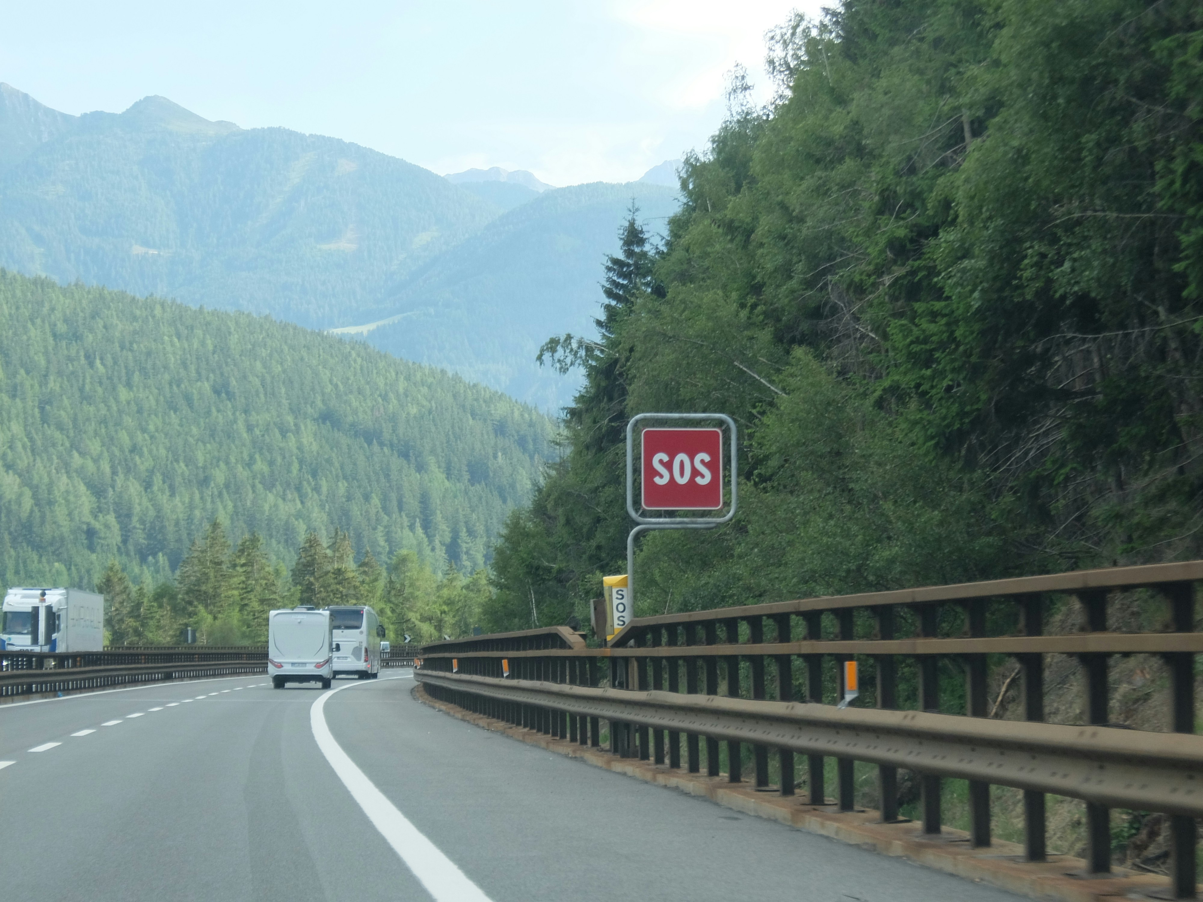 Curved highway with a red SOS sign rising over a wooden guardrail, mountains and forest in the background; road vehicles travel toward the distance.