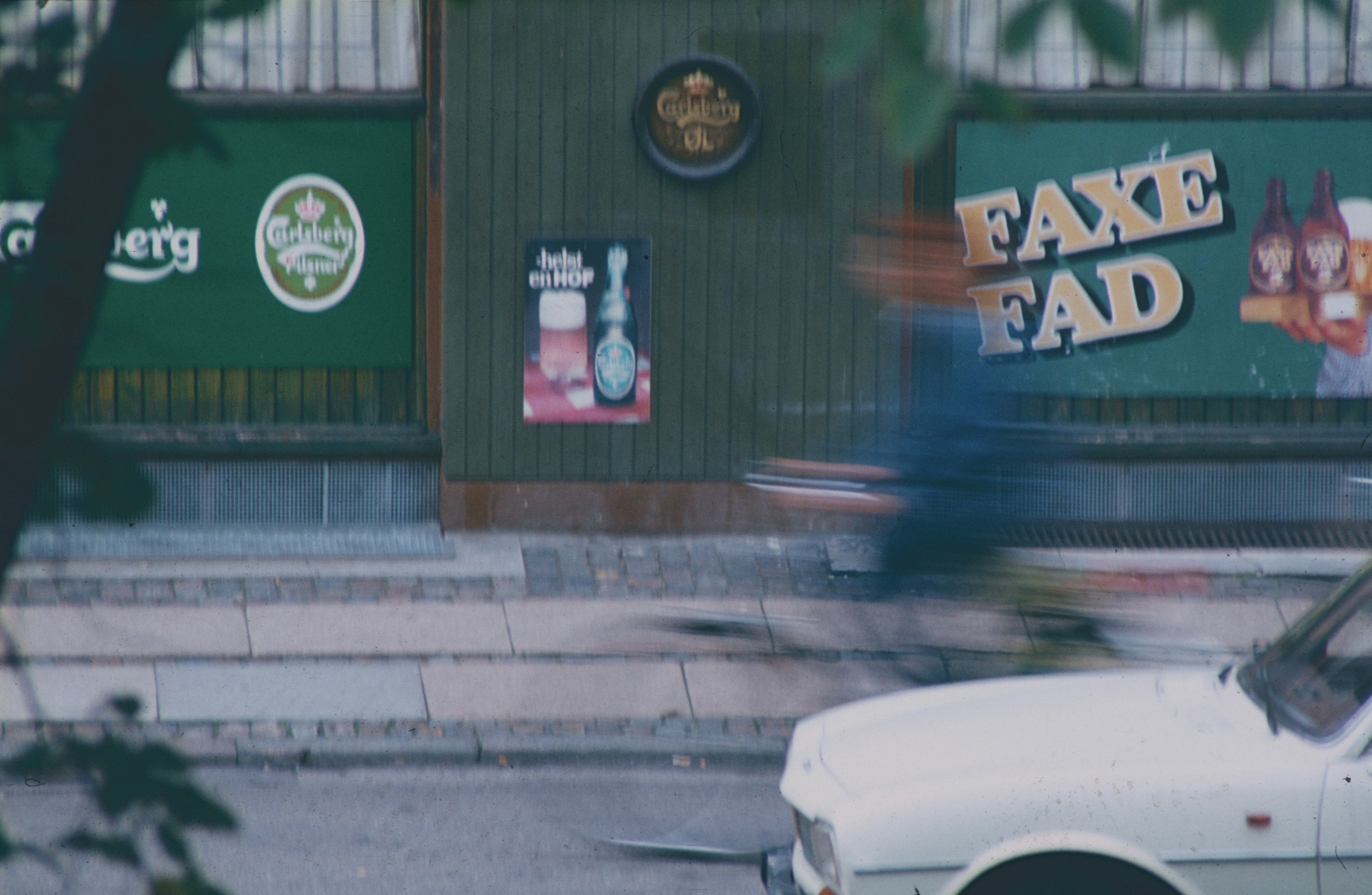 A candid color film photograph of a Danish pub facade in 1980s Copenhagen, plastered with bold beer advertising. Carlsberg Pilsner and Faxe Fad signage dominate the dark green frontage, alongside a classic "Helst en Hof" poster — iconic Danish beer brands of the era. A cyclist passes in a dramatic motion blur in the foreground, while a white vintage car is parked below. Glimpsed through tree foliage, the image captures an authentically nostalgic slice of everyday Copenhagen street life.