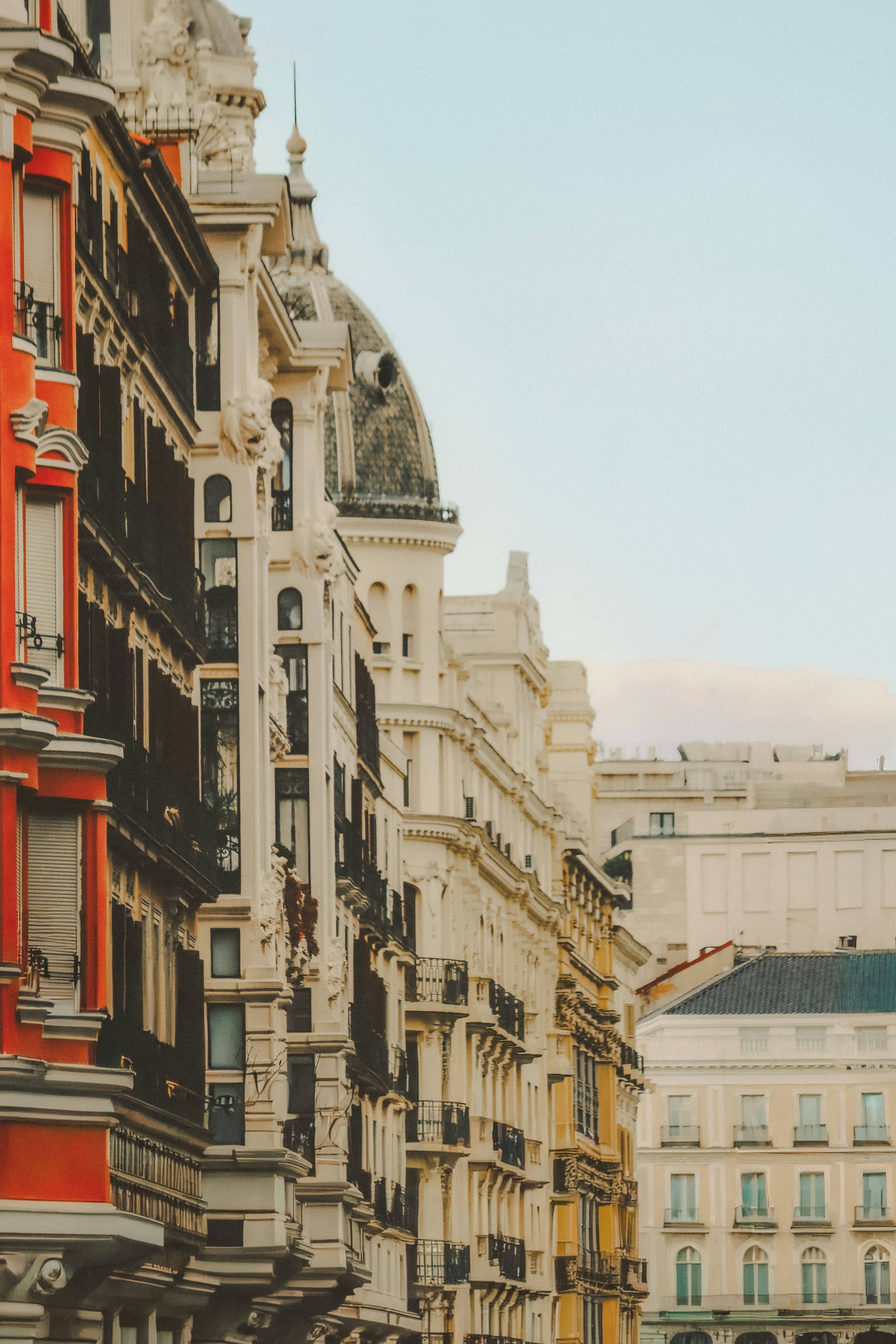 Row of ornate city façades lines the street, captured in soft light with prominent vertical architecture and a pale blue sky above.