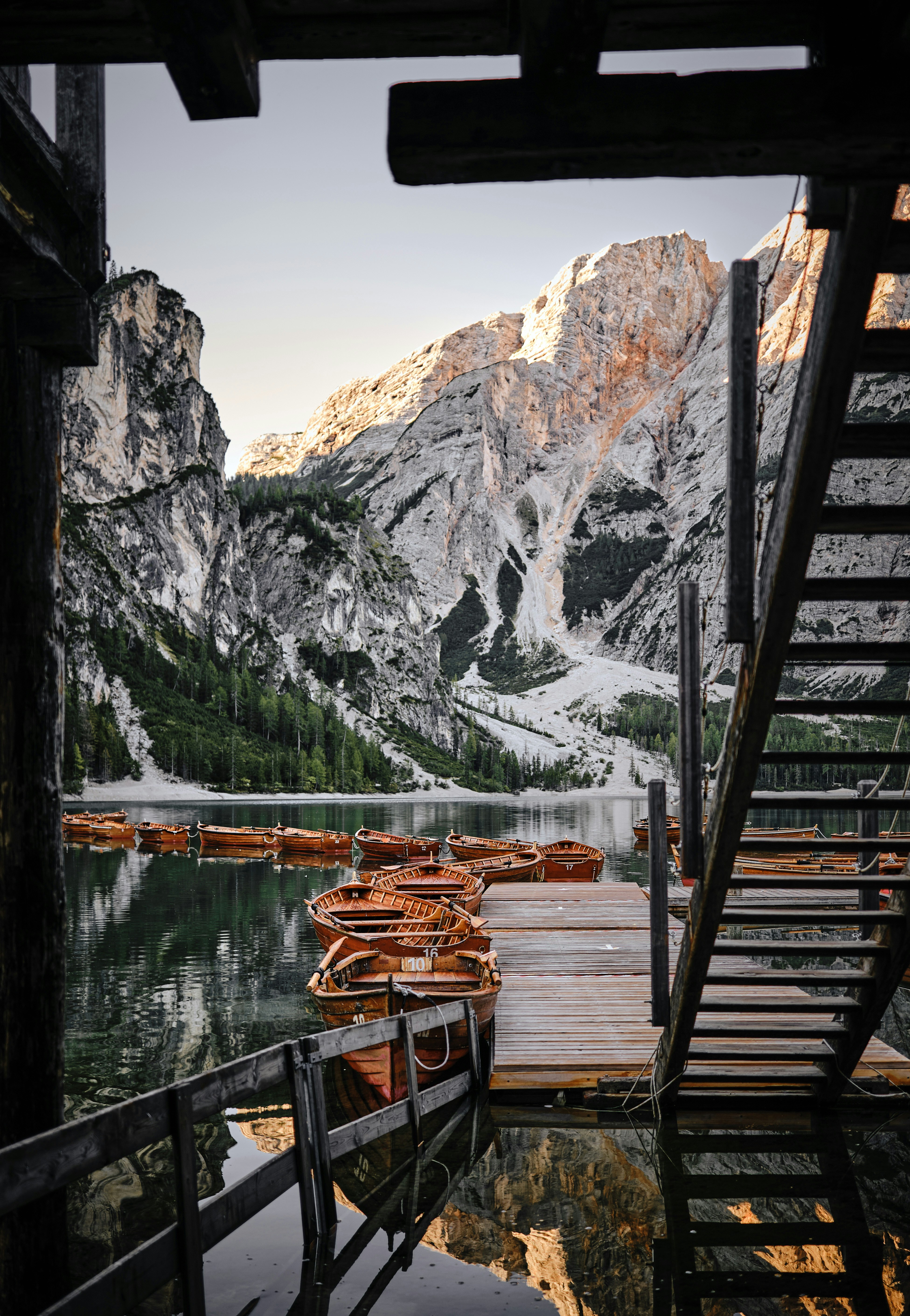Wooden stairs and dock framing lead to a calm alpine lake with rowboats and towering limestone peaks in the background. A composed landscape scene featuring reflections on still water.