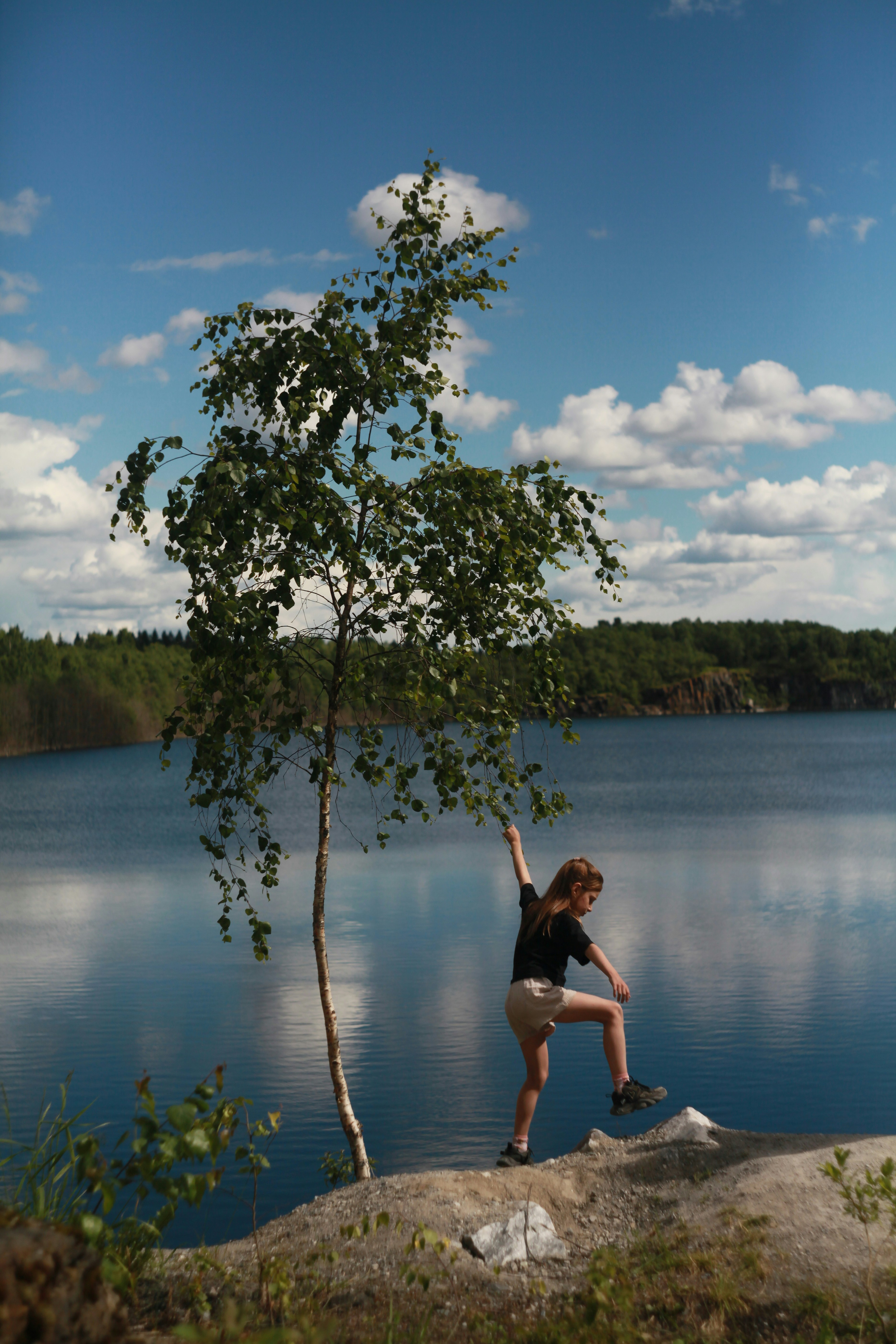 A girl steps confidently onto a rocky ledge beside a calm lake with a lone tree in the foreground; the scene captures a moment of balance and exploration by the water's edge.