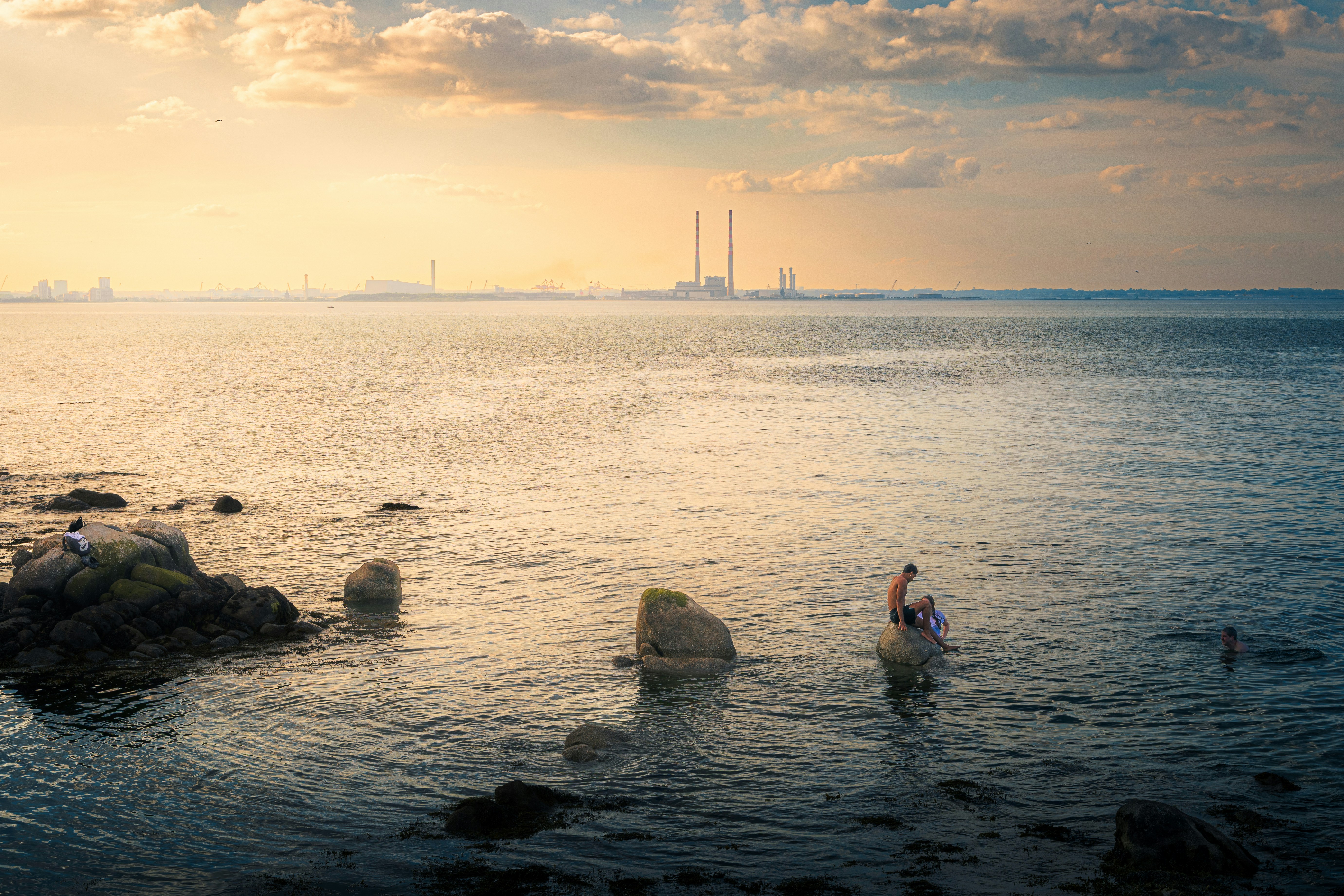 Sunlit low tide: distant factory skyline meets quiet shoreline and swimmers