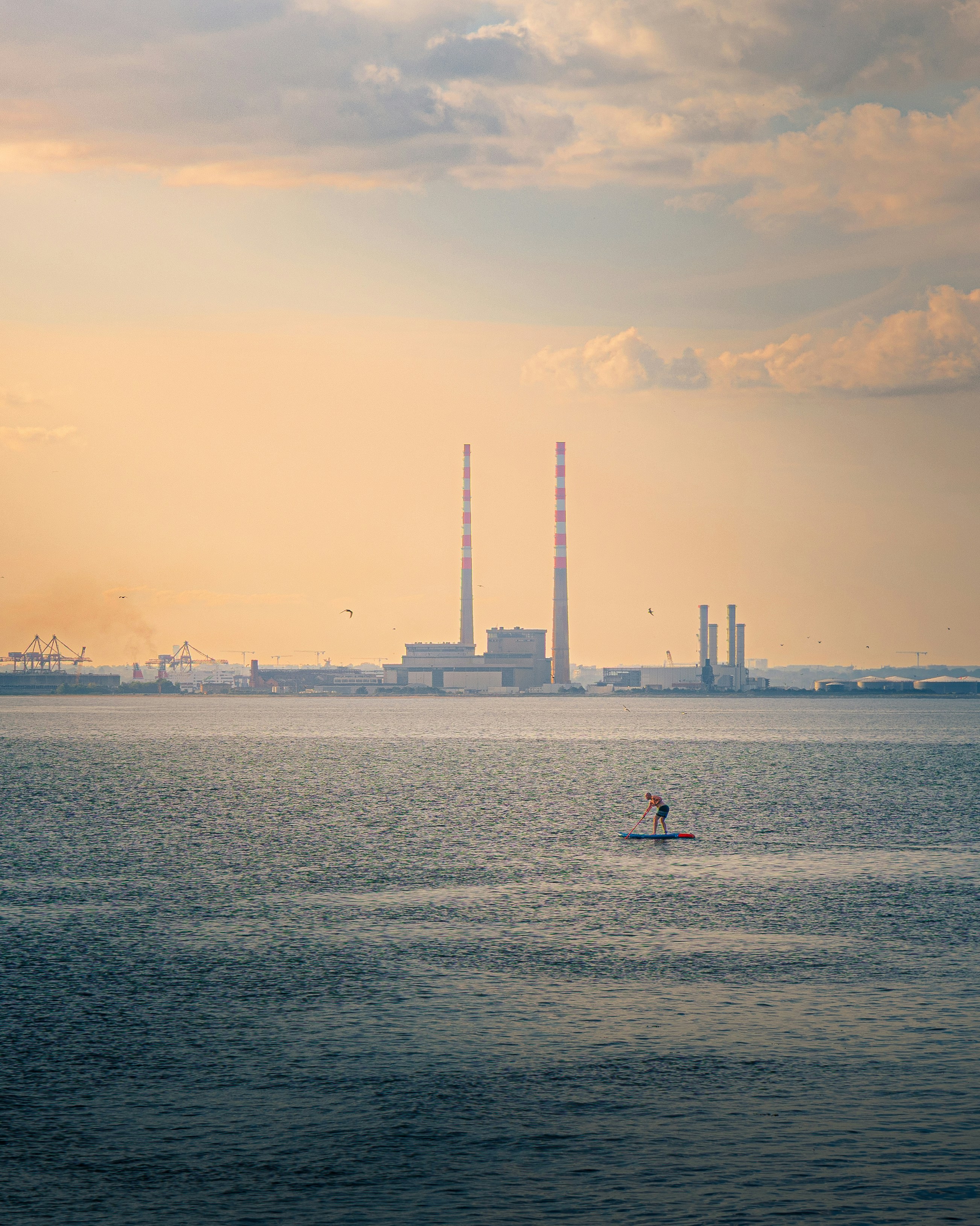 Sunset Glide: A lone surfer on a tranquil estuary under industrial silhouettes