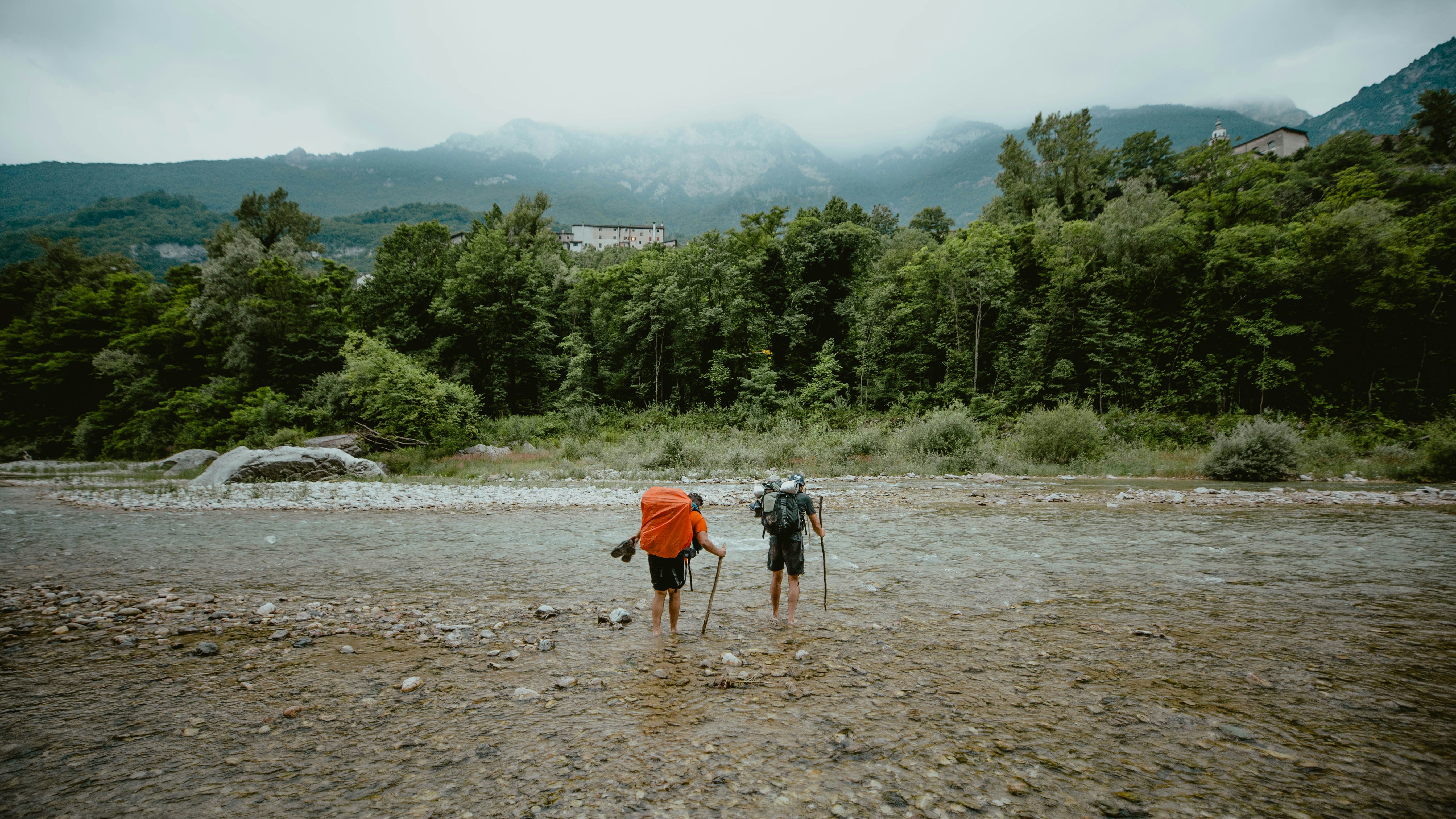 Two Wanderers Crossing a Pebble River under Mountain Canopy
