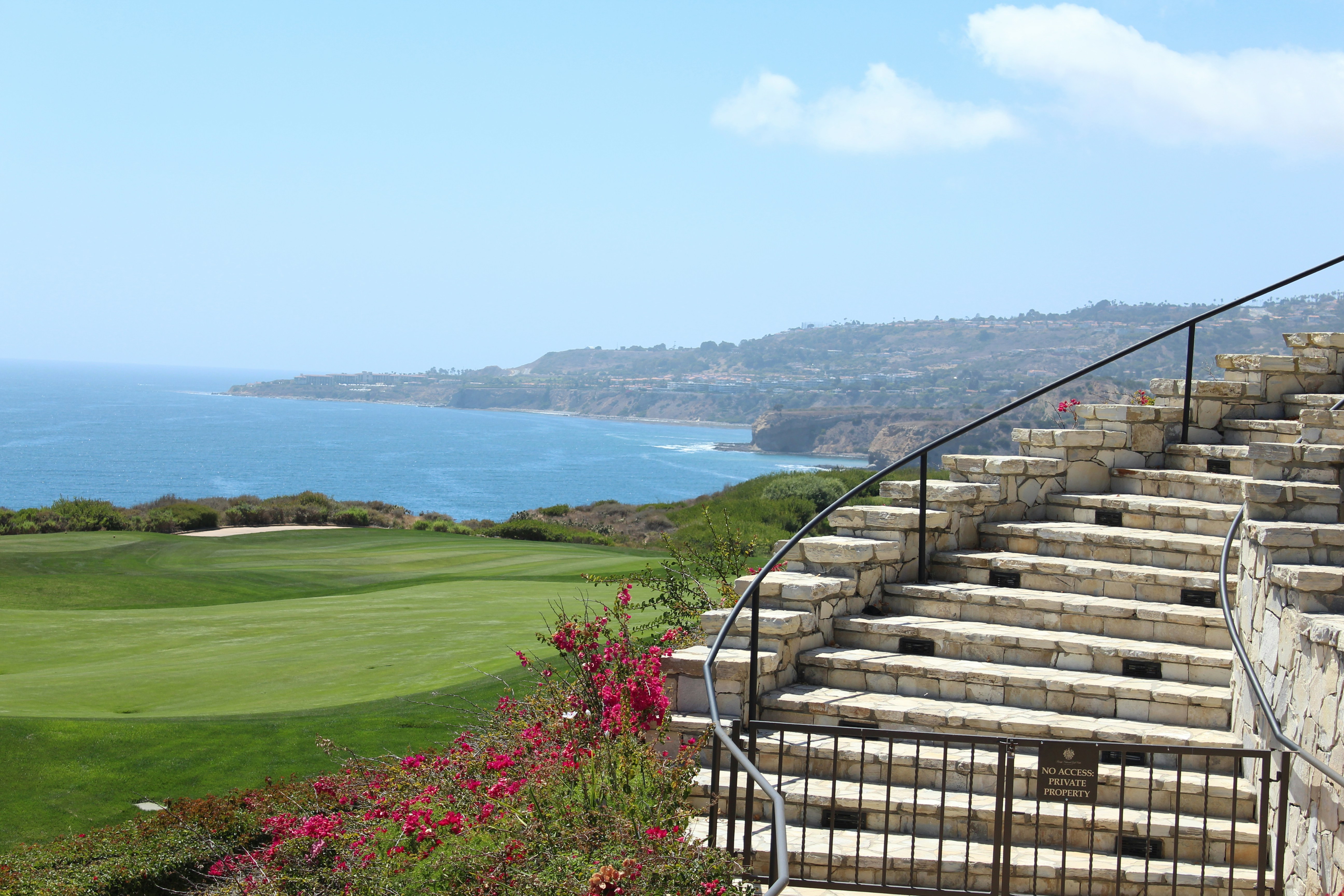 Coastal golf course with terraced stone steps along a railing, green fairway to the left and distant cliffs by the sea.