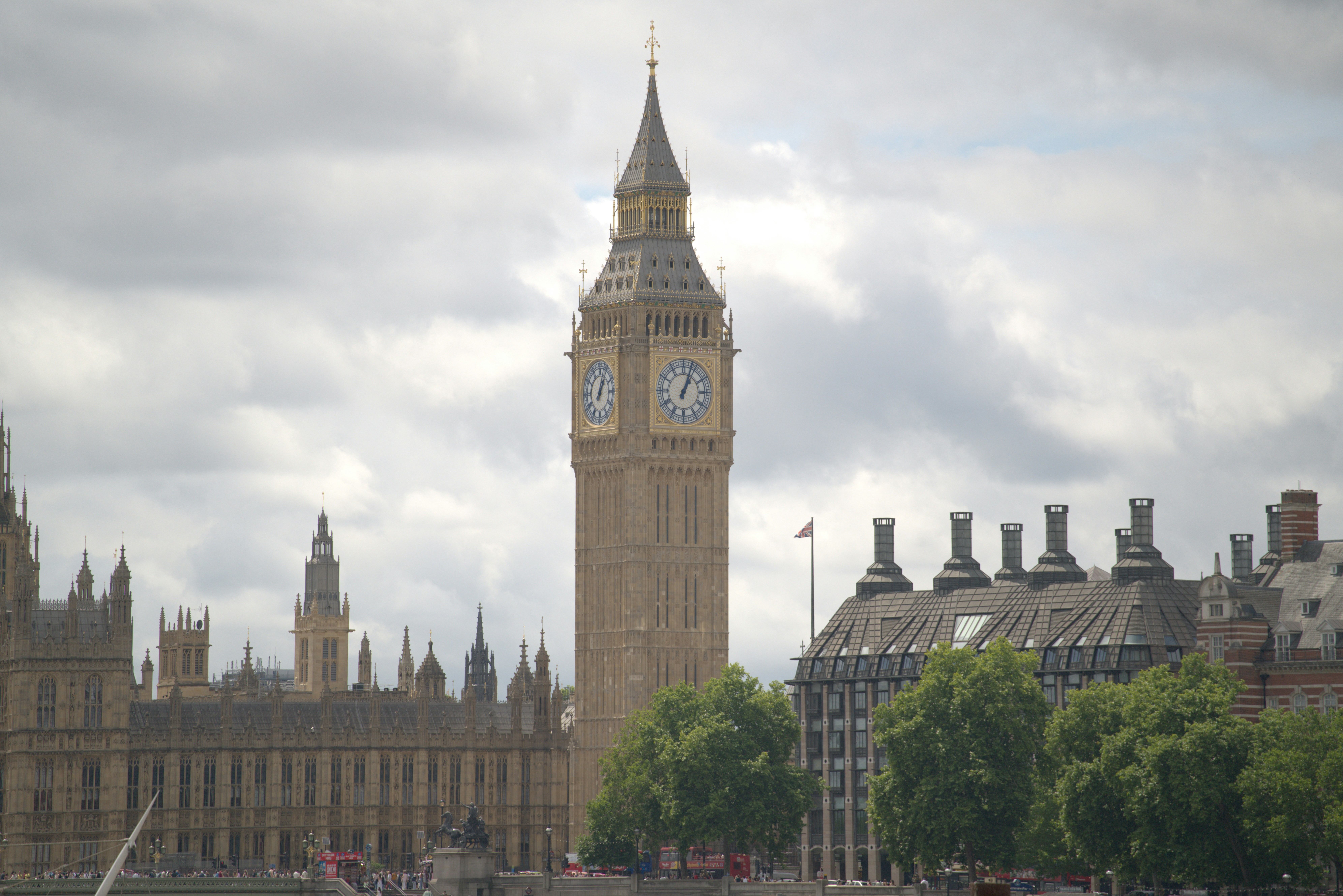 Towering clock tower of the Elizabeth Tower with adjacent Parliament buildings, riverfront trees, and a cloudy sky in London. A documentary street-scene capturing historic architecture and a calm civic backdrop.