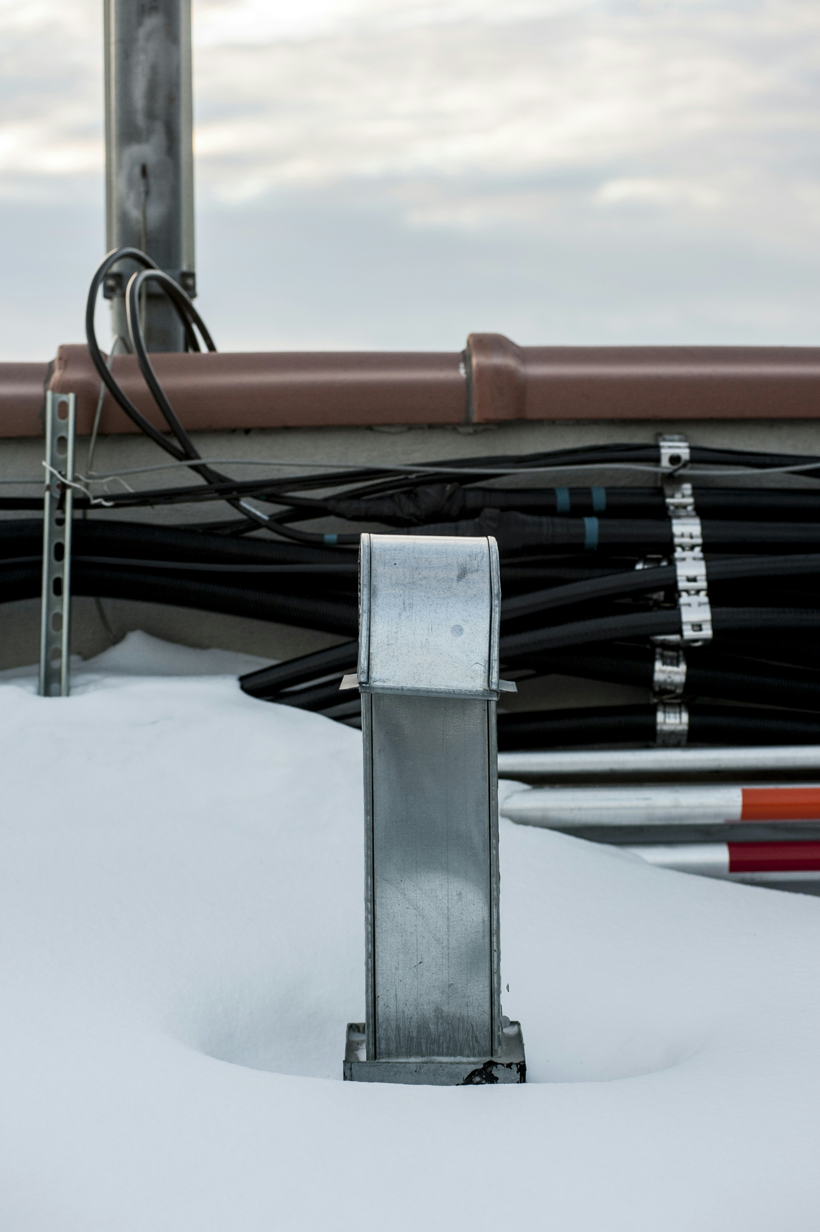 A close-up of a metallic rooftop fixture emerging from snow with bundled cables and piping in the background, illustrating industrial rooftop details. This is a photograph.