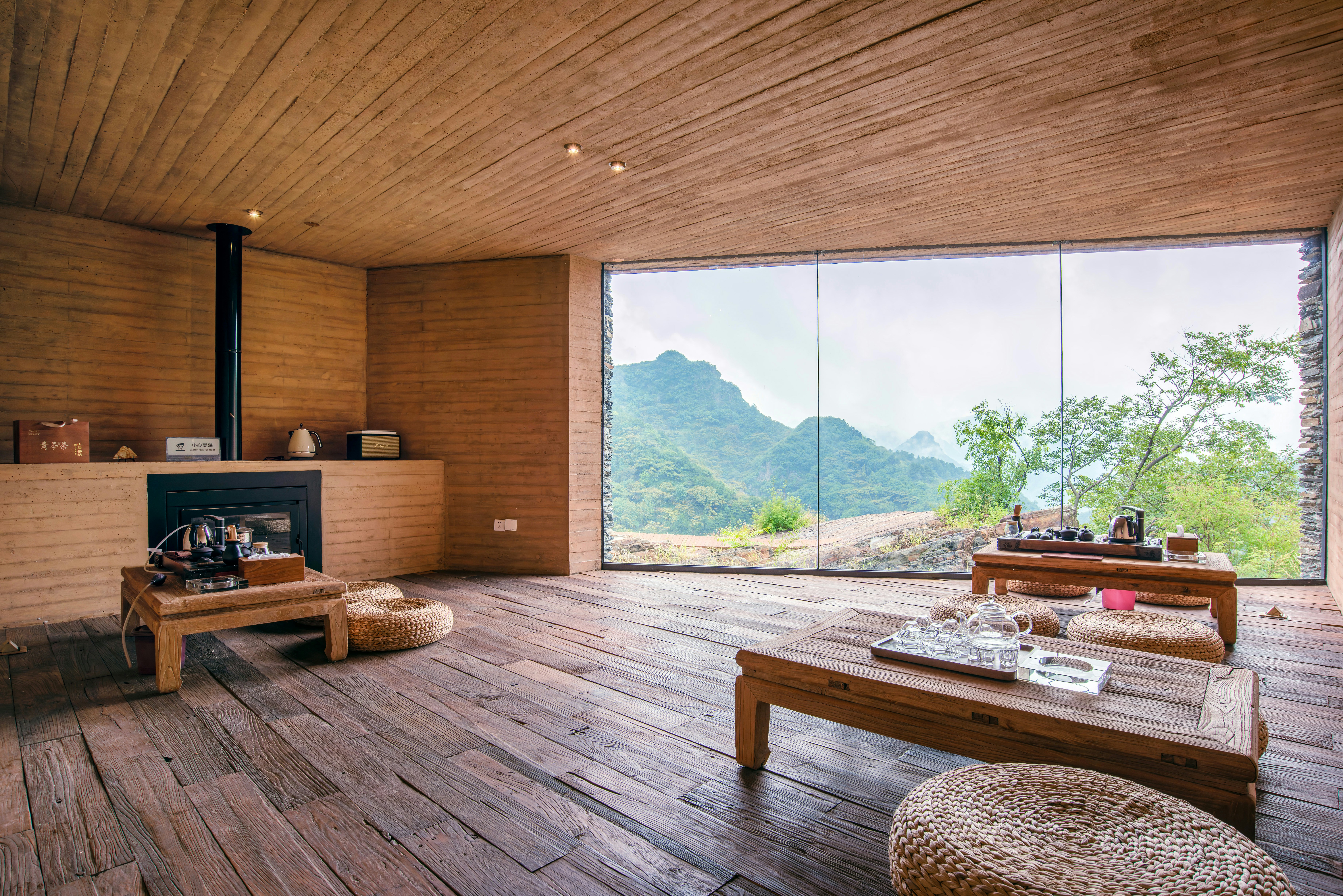 Bright, open living space with wooden floors and ceiling, large glass wall revealing lush mountains outside; a low wooden coffee table and woven seating add a minimalist, calming aesthetic.