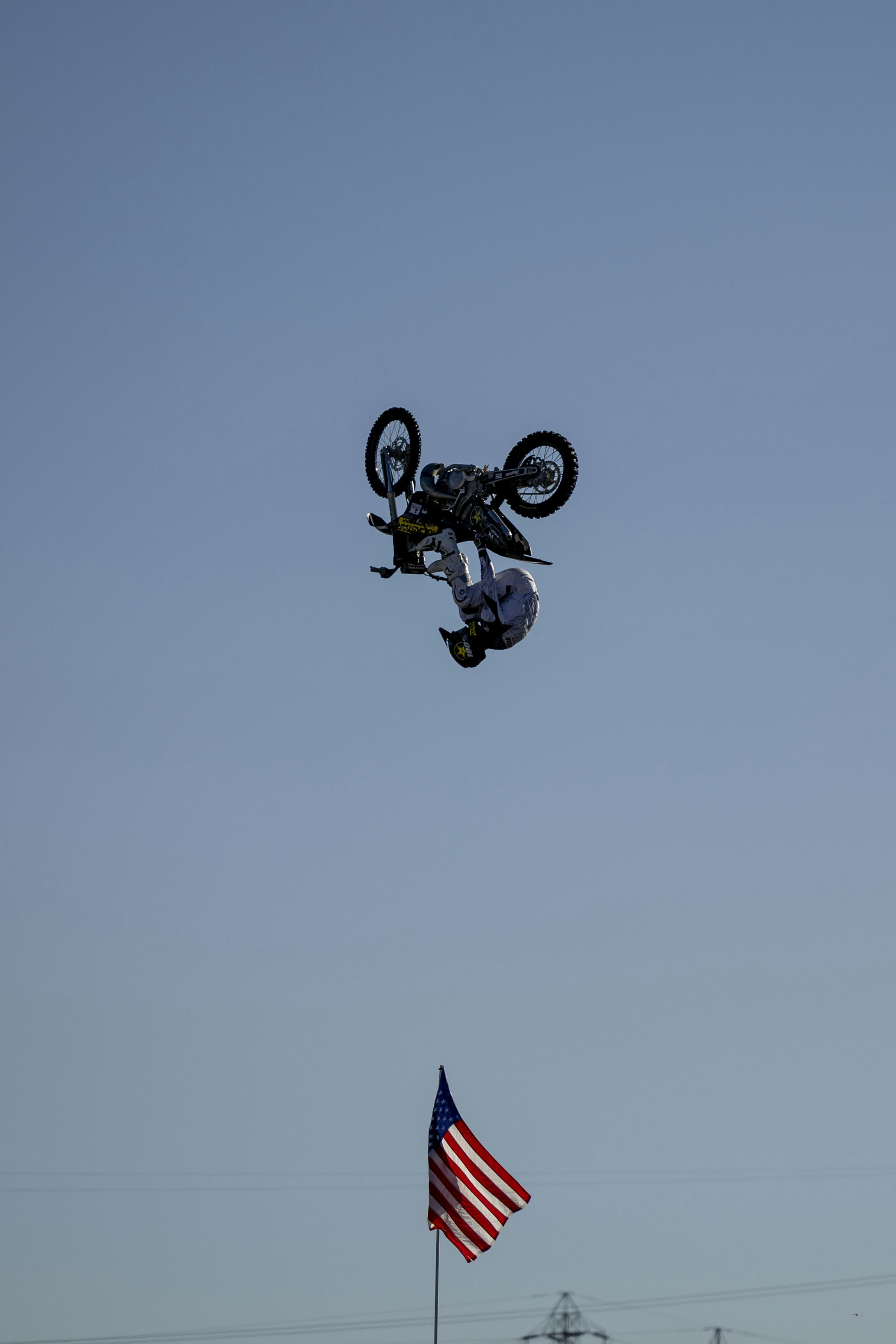 Motocross rider completes a mid-air flip against a pale blue sky with an American flag visible toward the bottom. The shot emphasizes the stunt and empty space around, highlighting the isolation of the airborne rider.
