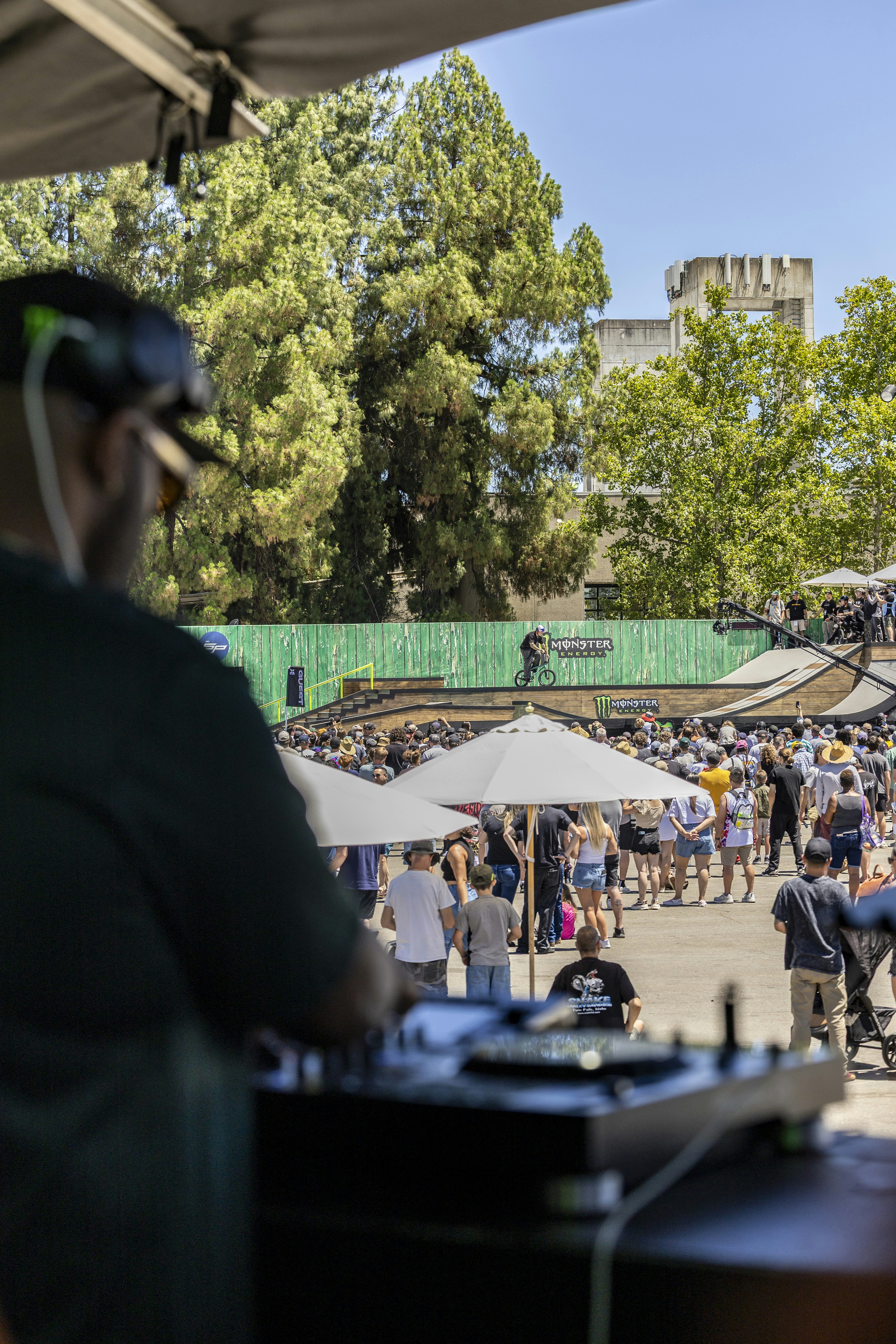 DJ booth in foreground with a bustling outdoor skate event crowd and green barriers in background; tents and umbrellas dot the plaza under a bright day.