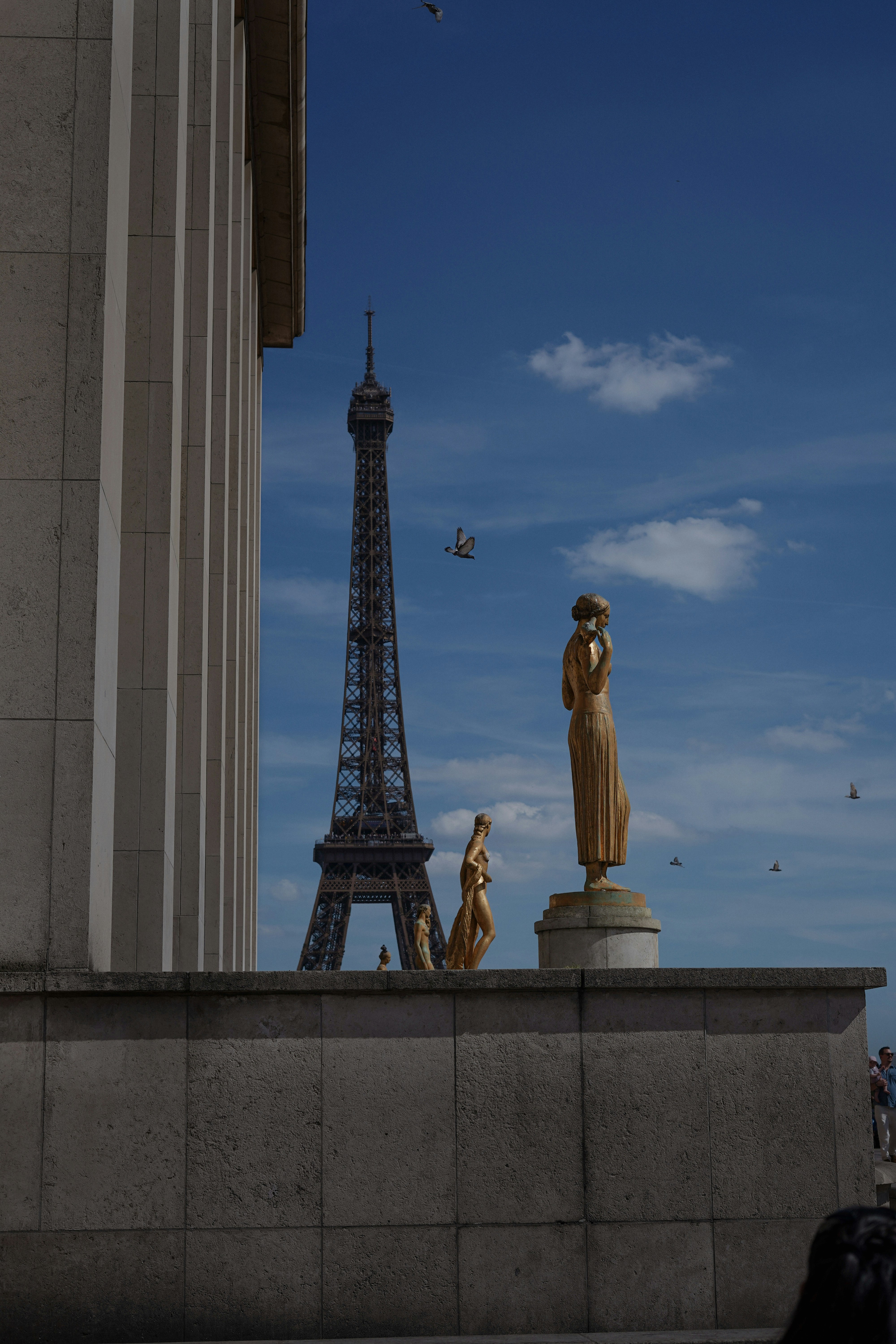 Golden statues stand on a stone ledge with the Eiffel Tower and a blue sky in the background.