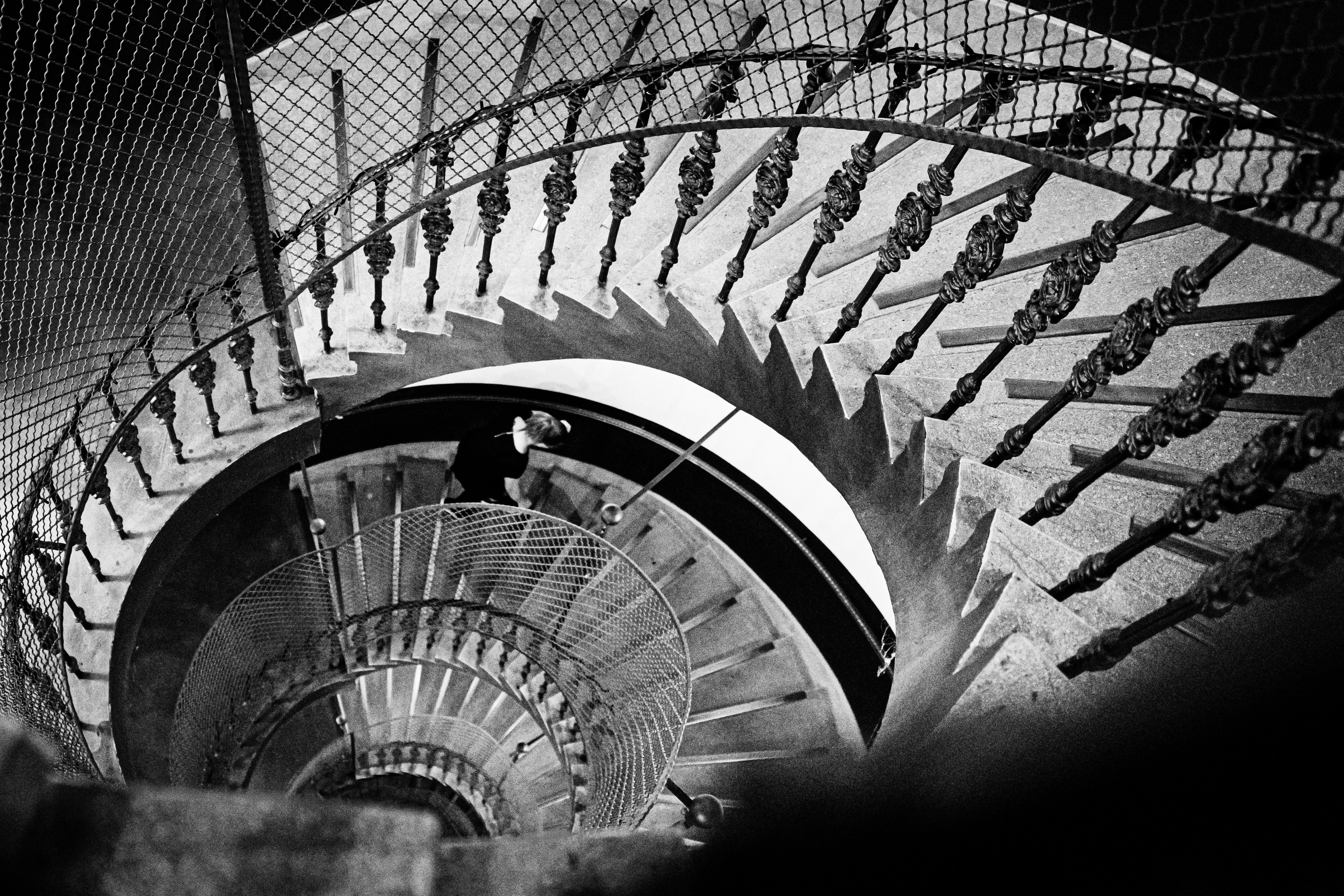 Winding iron staircase seen from above, monochrome tones emphasize texture and pattern; the image focuses on the spiraling rails and steps as they recede into the lightless center.