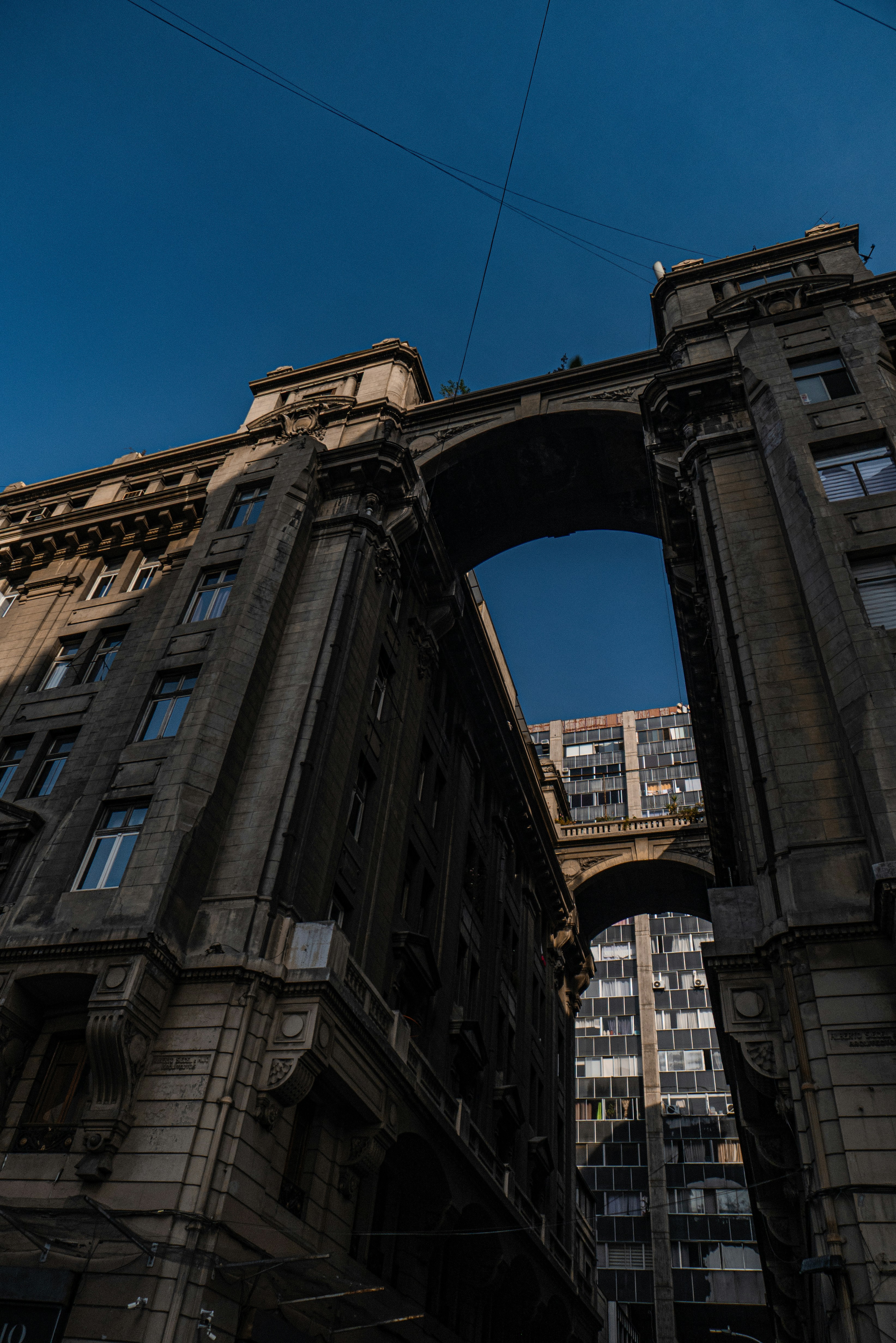 Stone archways frame a narrow urban gap with a modern building peeking through; deep shadows emphasize texture and scale. This is a cityscape photograph capturing a dramatic architectural passage.