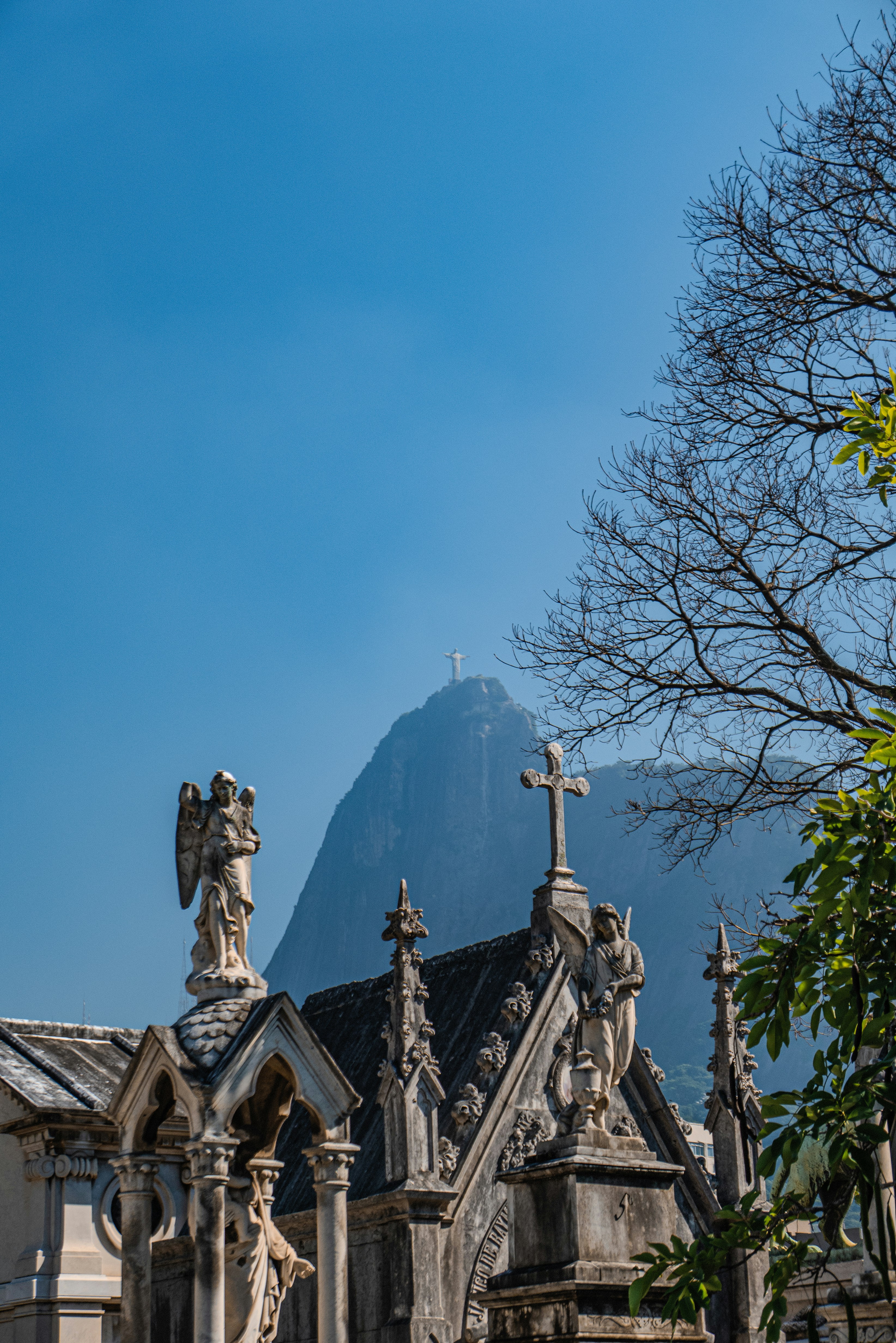Bronze-gray cemetery statues and ornate Gothic spires in the foreground with a distant mountain peak in the background under a clear blue sky.
