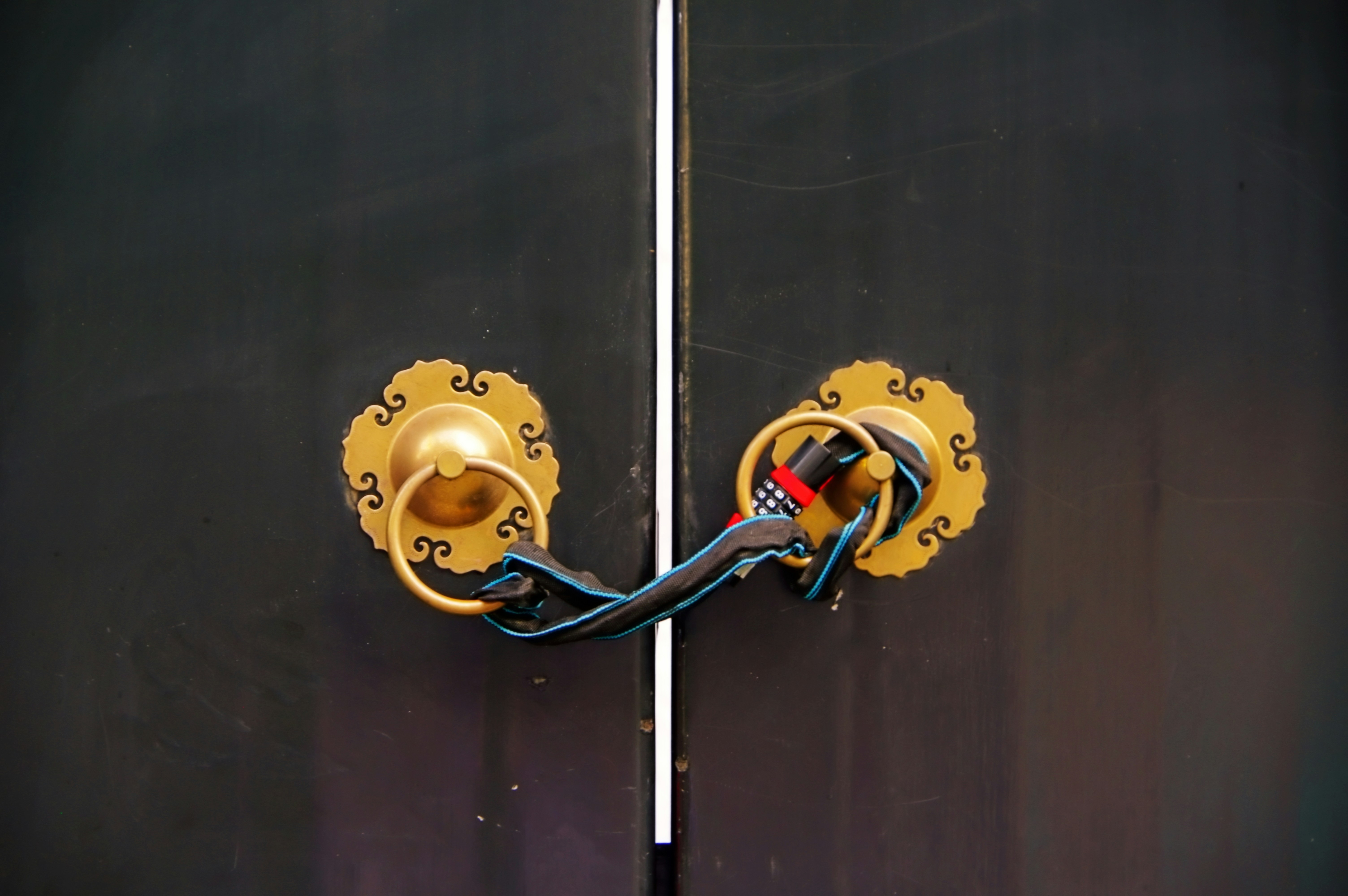Two ornate door handles with yellow-gold rosettes on a dark door, linked by a padlock and chain; the contrast highlights the locked pairing and the subtle vertical gap between doors.