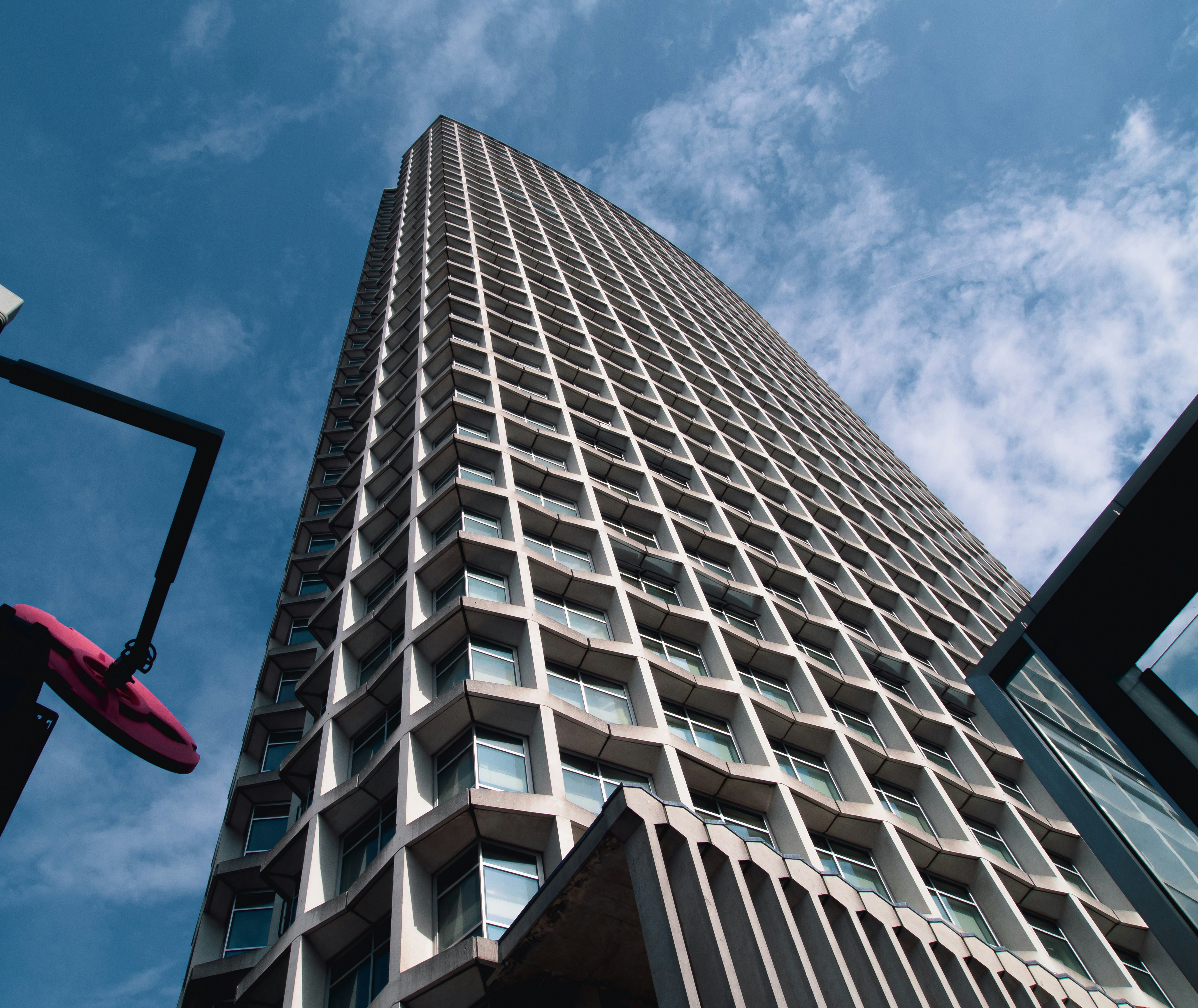 Tall concrete-and-glass skyscraper viewed from a low angle with a blue sky; a modern urban structure dominates the frame and a pink streetlamp hints at surroundings. Structural grid and reflections define the scene.