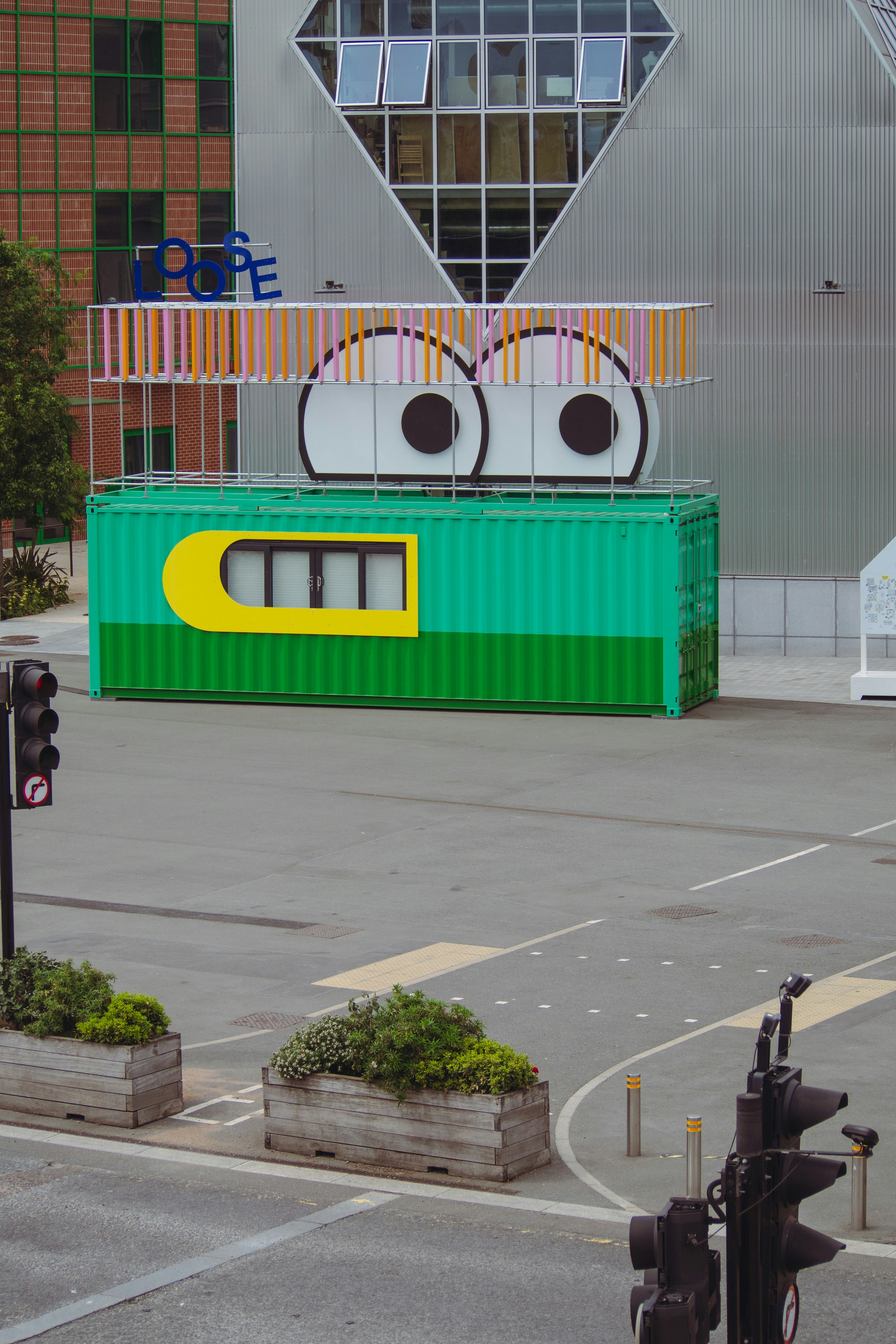 Bright green storage container with bold yellow and cut-out shapes sits in an open plaza, topped by a playful sculpture with large cartoon-like eyes and striped railing. The scene includes surrounding planters and a modern building facade.