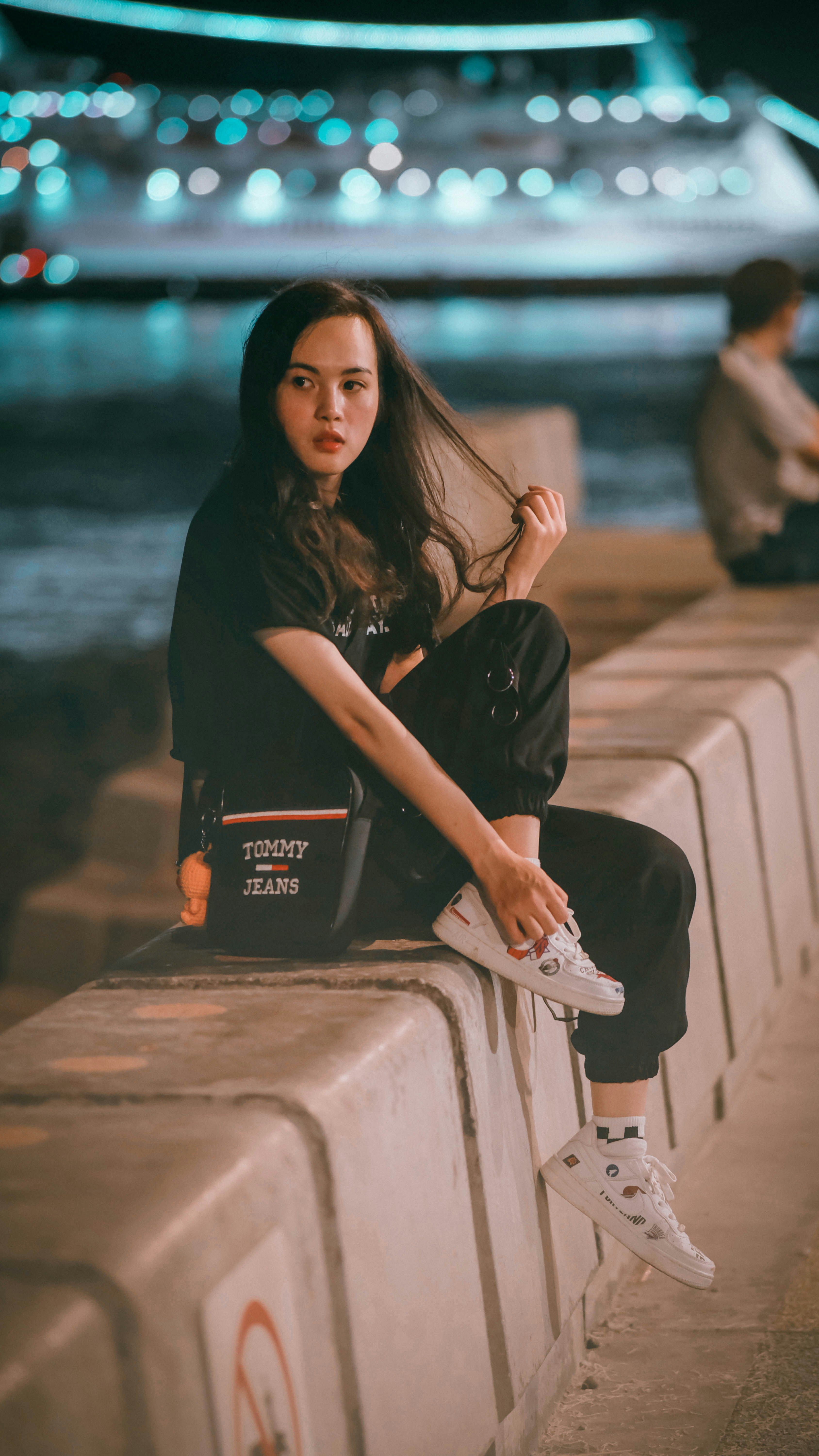 Young woman sits on a concrete seawall by the waterfront at night, catching the breeze in her hair with a small crossbody bag and white sneakers. A blurred skyline and another figure in the background set a moody, urban scene.