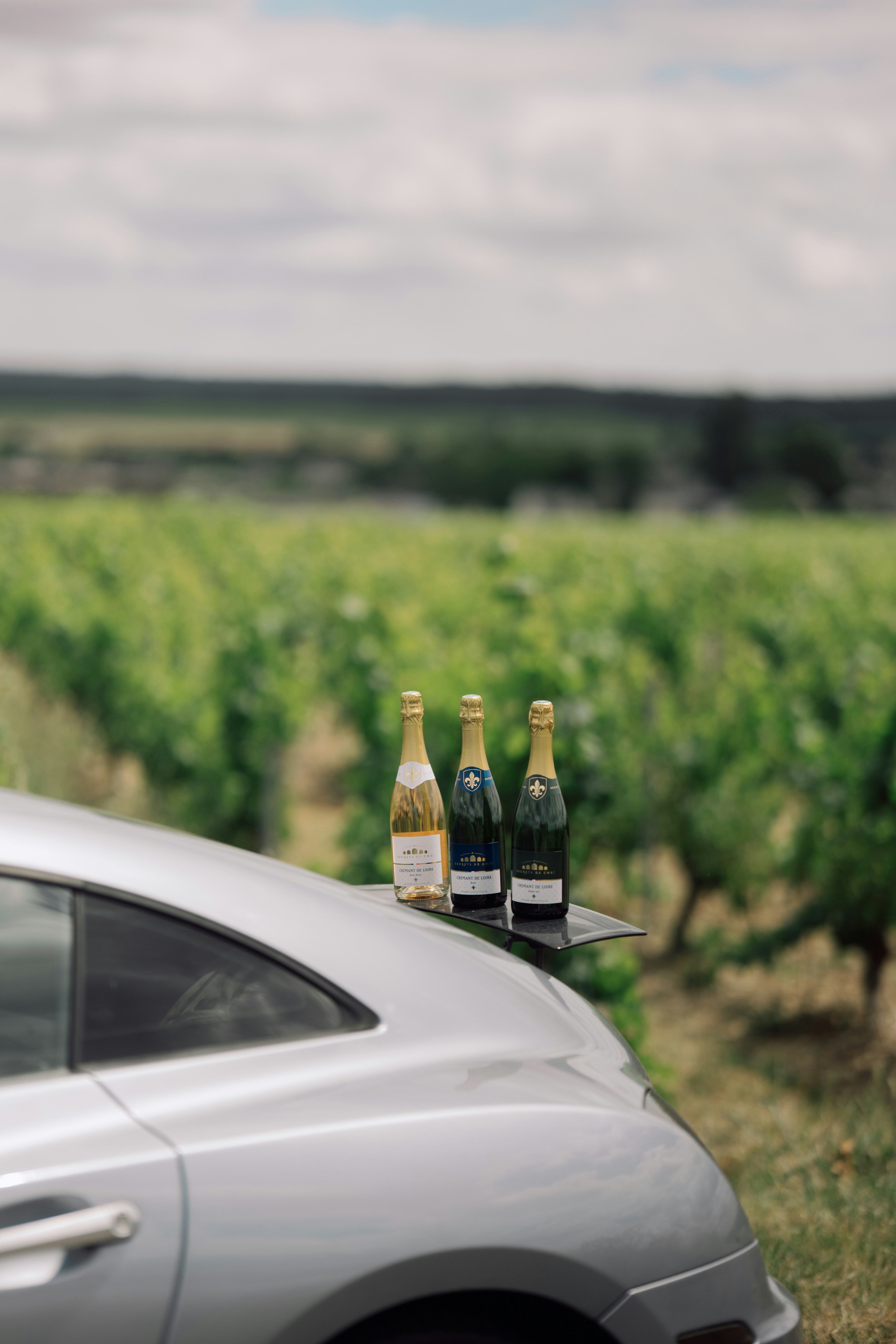Three sparkling wine bottles sit on a narrow tray atop the rear edge of a silver car, with rows of vines fading into the distance and a cloudy sky above.